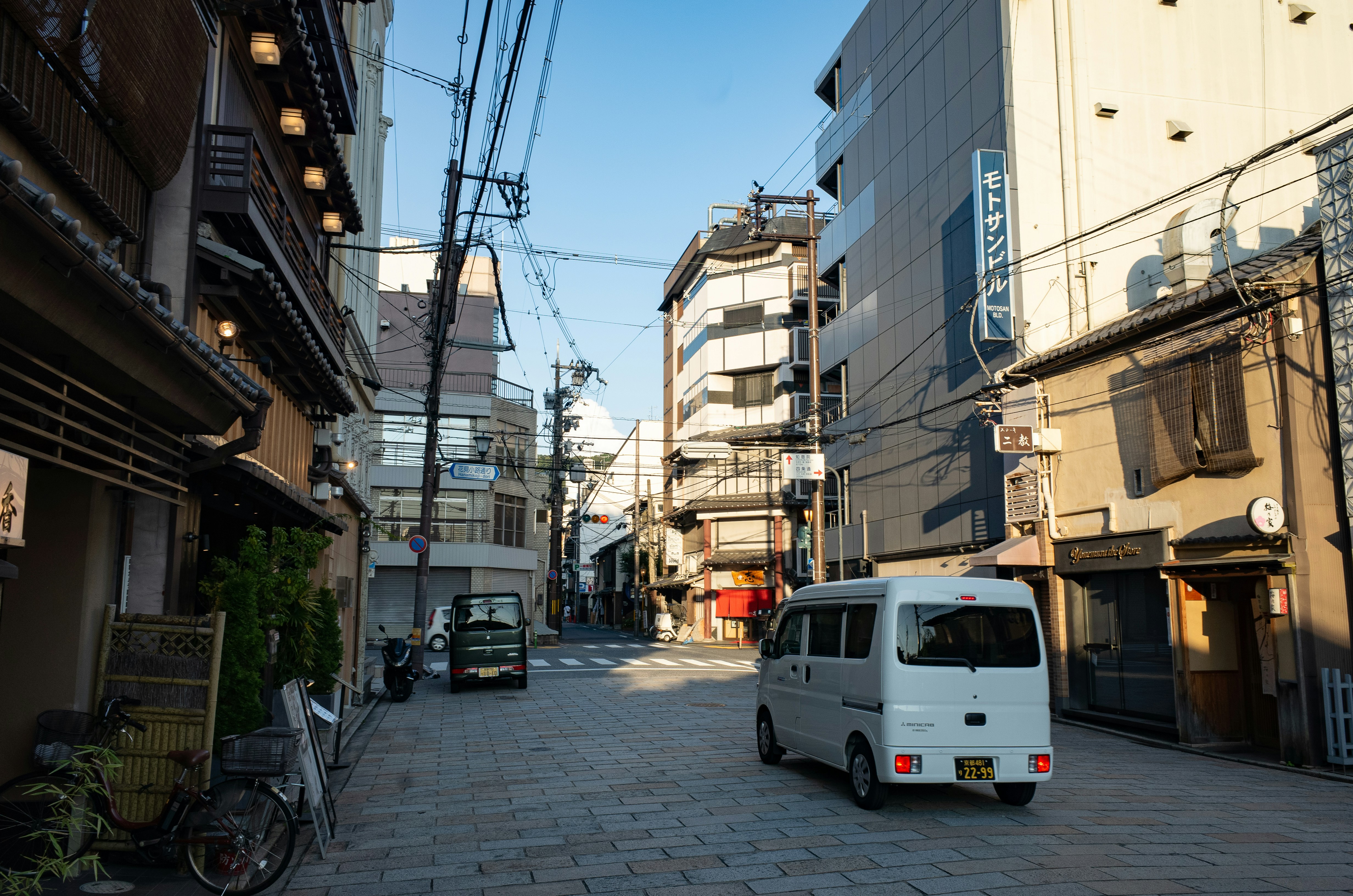 Narrow urban street lined with low-rise buildings and parked vans; a quiet morning scene with clear sky and overhead utility lines.