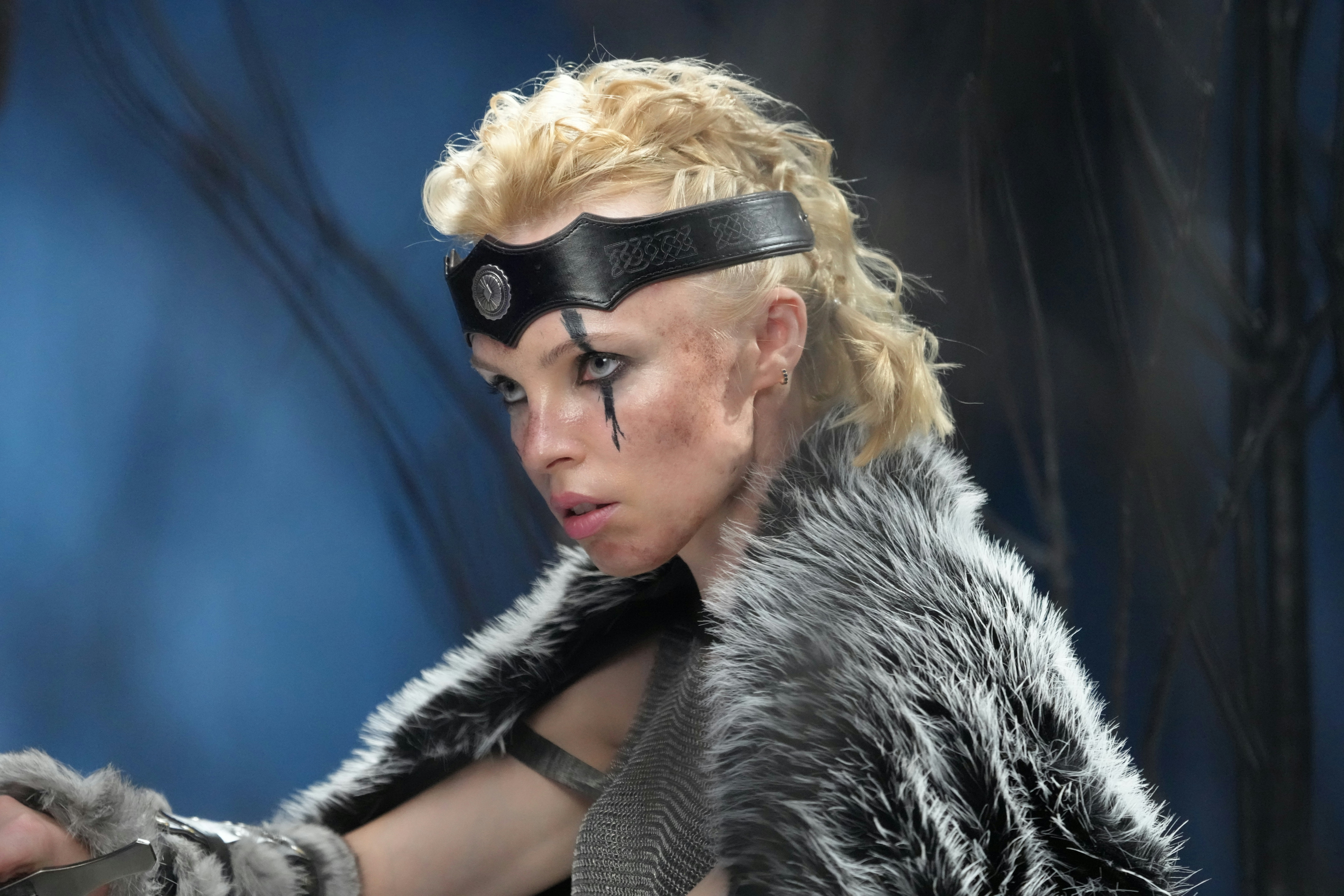 Close-up of a battle-ready character with blonde hair, fur cloak, and a leather headband, eyes intense and profile turned slightly downward, suggesting tension and readiness.