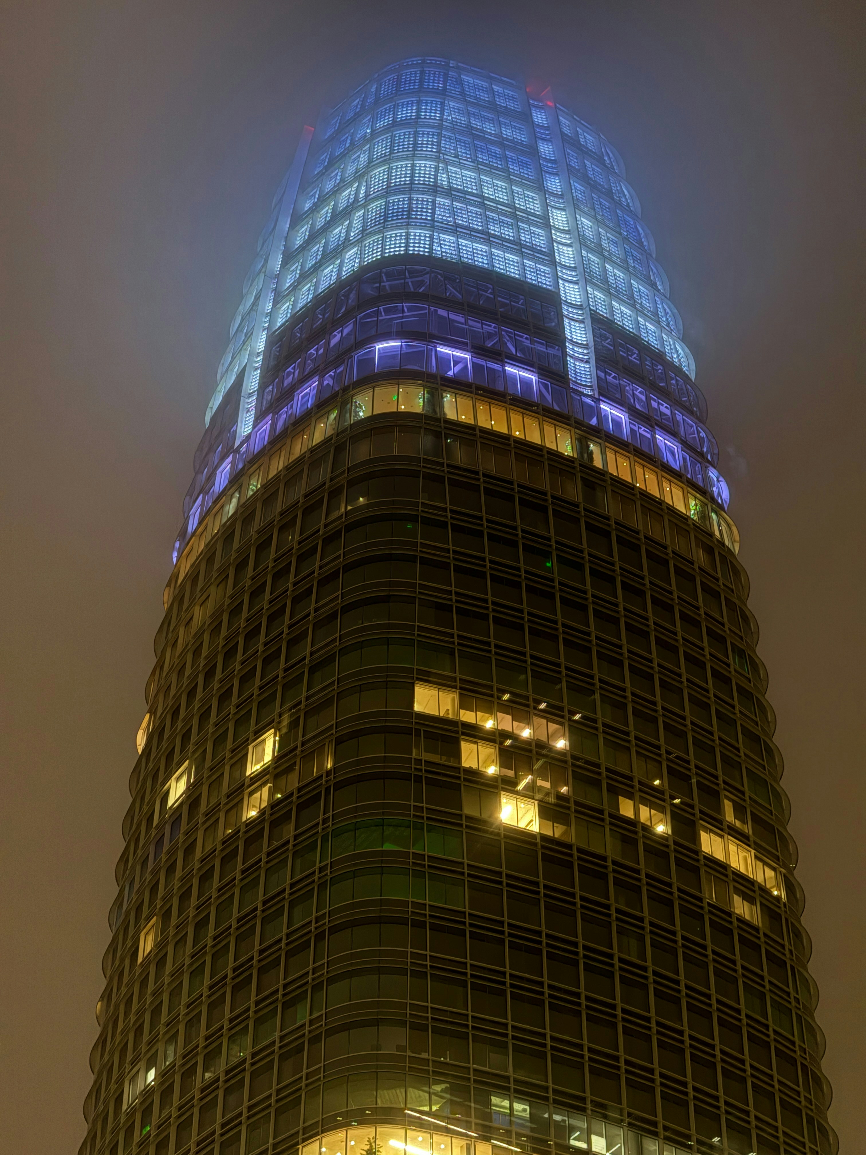 Tall skyscraper illuminated in blue and amber lights rises into a misty night sky, its glass facade radiating with layered horizontal bands.