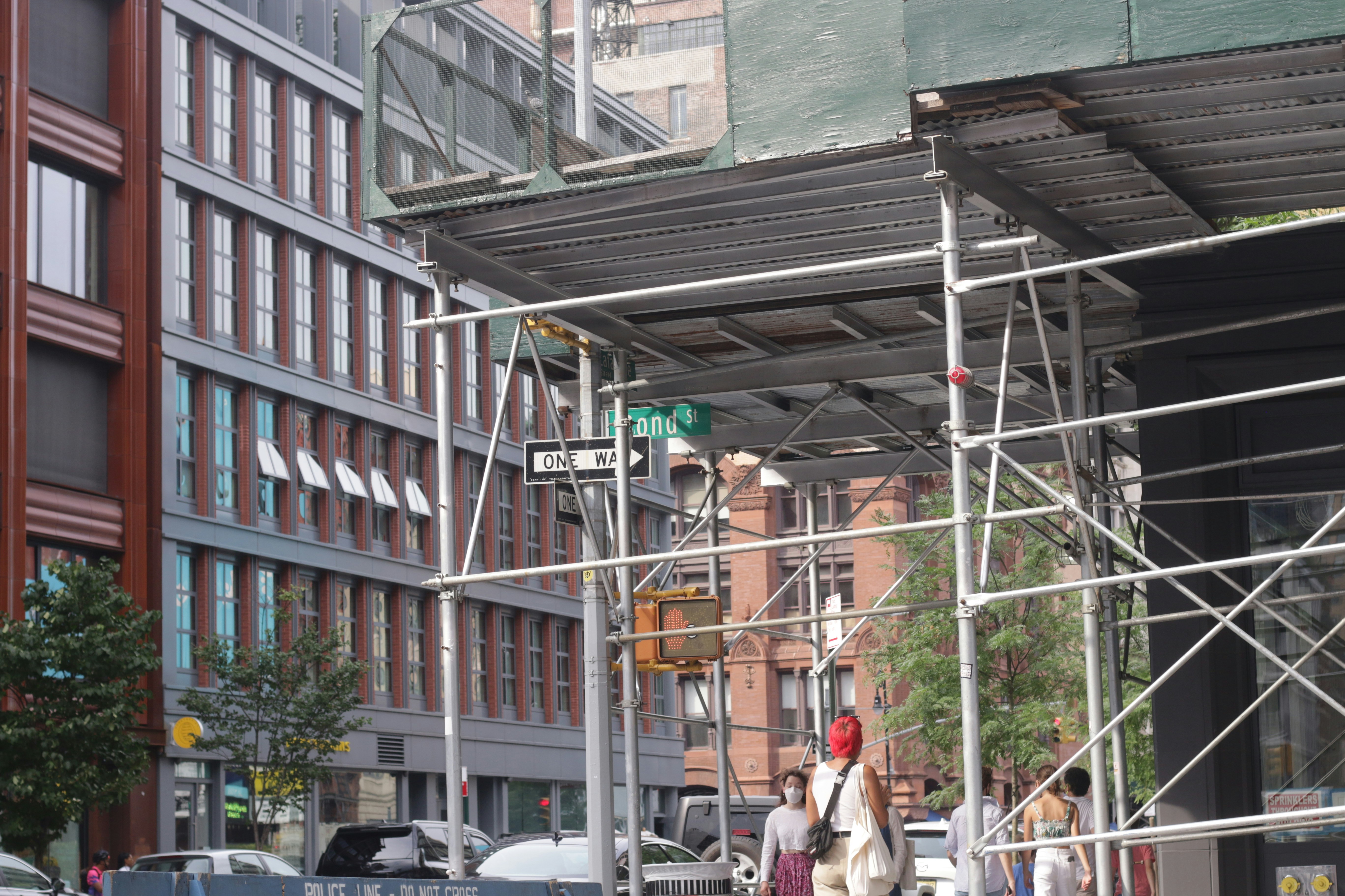 Street scene with scaffolding under construction alongside a row of modern buildings; pedestrians and vehicles present, conveying urban activity and construction context.
