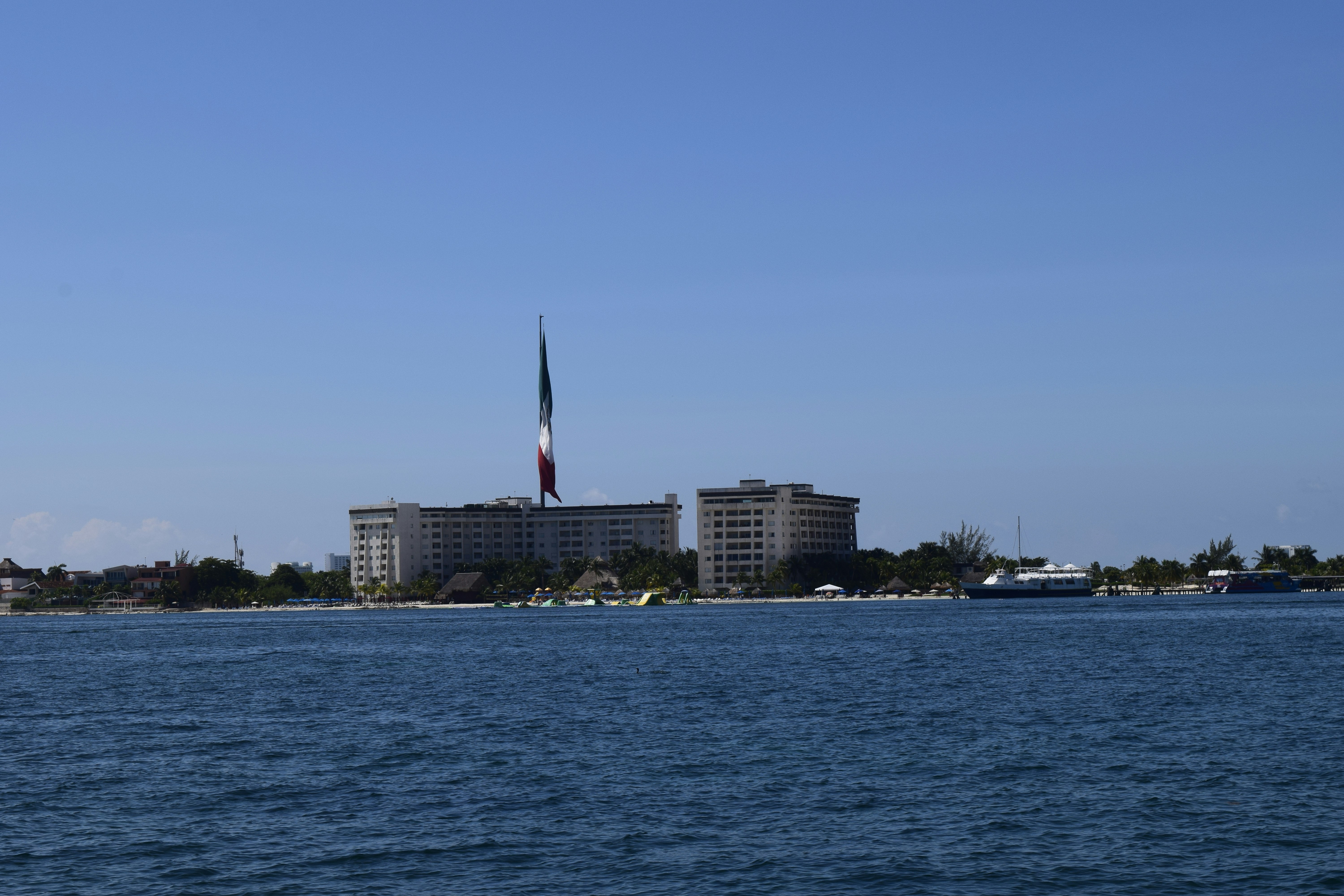 Coastal city skyline with a tall flagpole and a flag centered near the horizon, across a calm blue sea. This is a straightforward landscape shot featuring the shore buildings and open water.