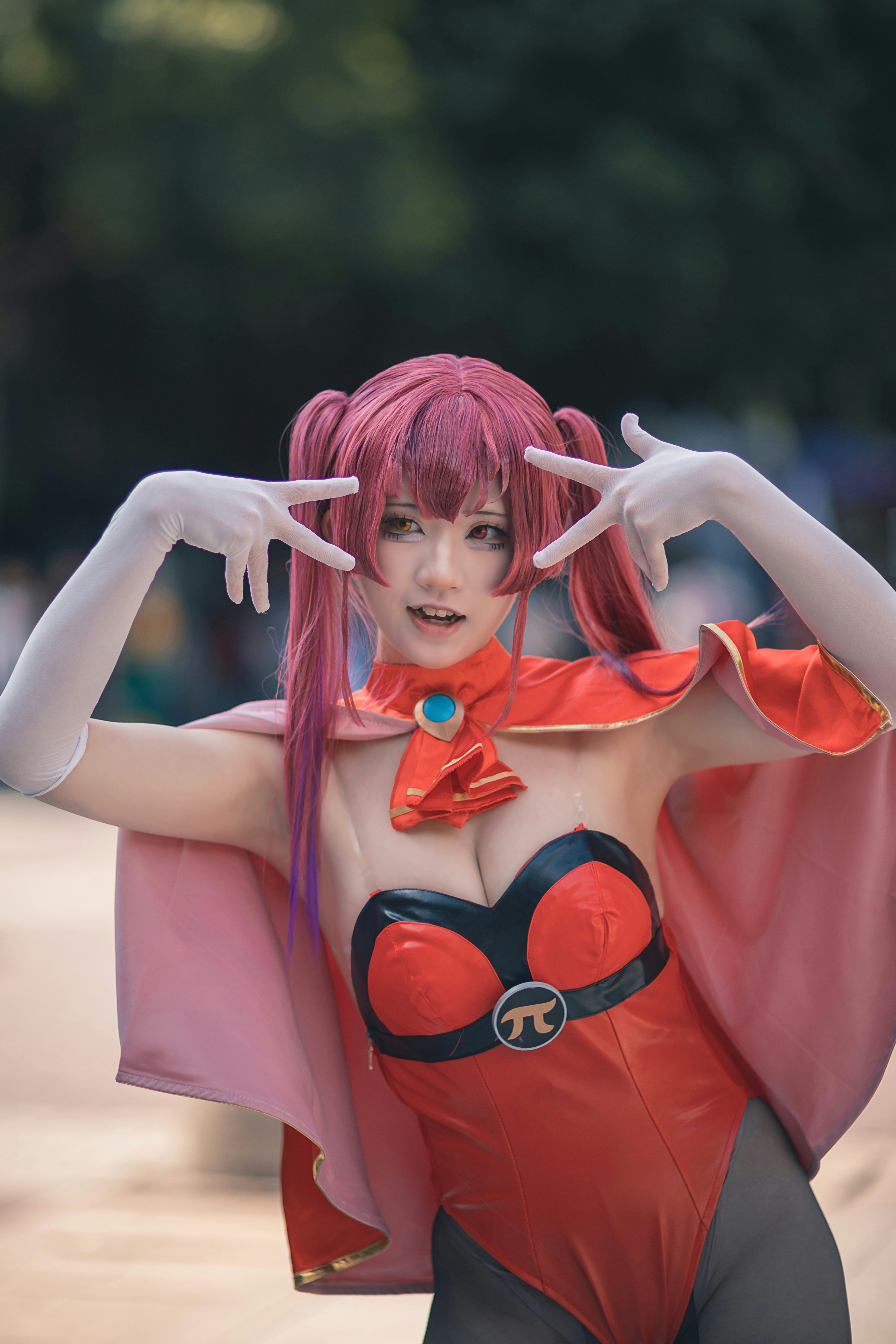 Character cosplayer posing with peace-sign gesture in front of a blurred outdoor backdrop; costume features red bodice with heart motif and pink cape, gloves, and twin ponytails. Indicates a convention or event setting.
