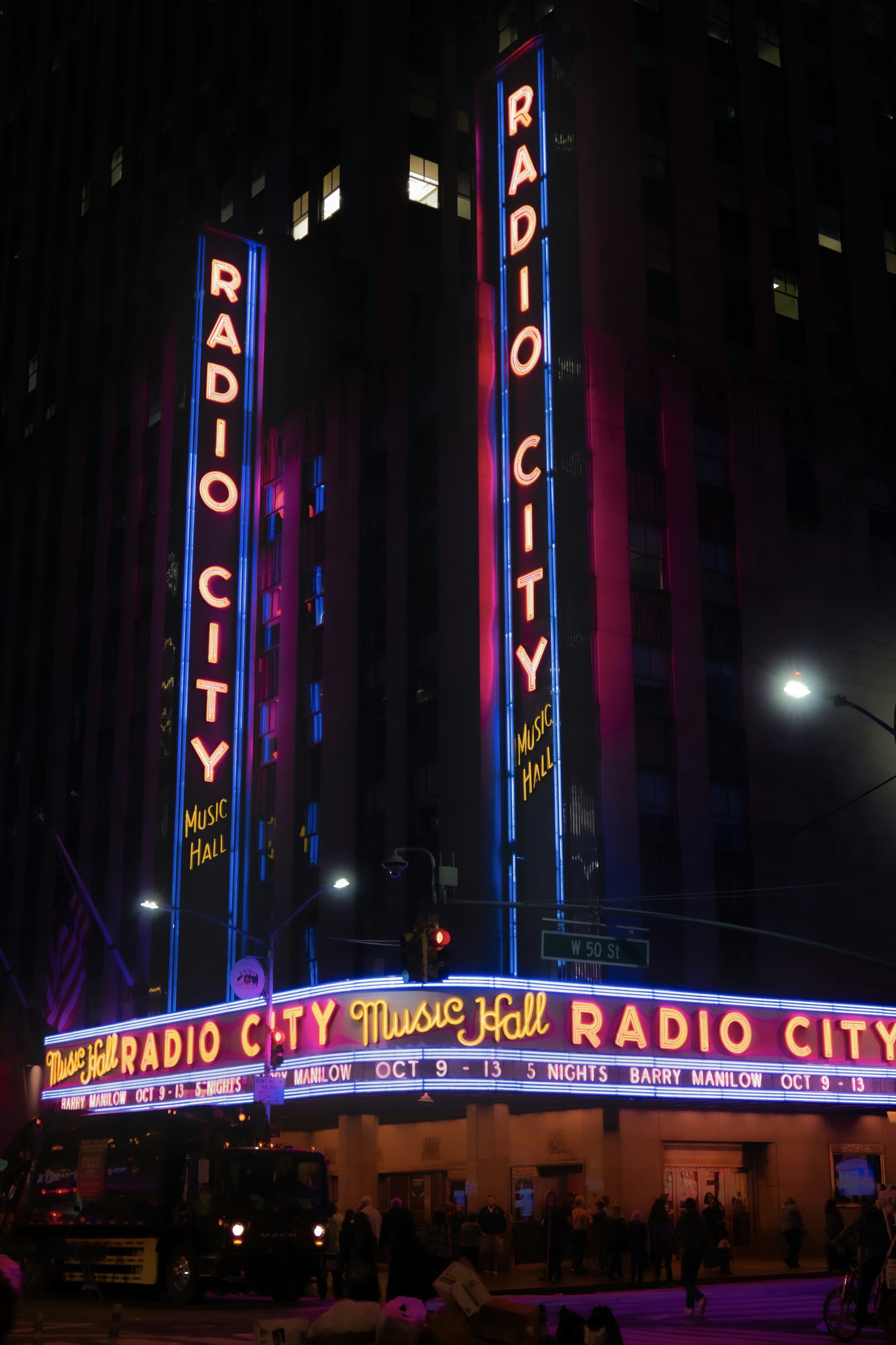 Neon-lit signs outline the marquee of Radio City Music Hall, with vertical signs spelling RADIO CITY towering above a bustling street scene at night.