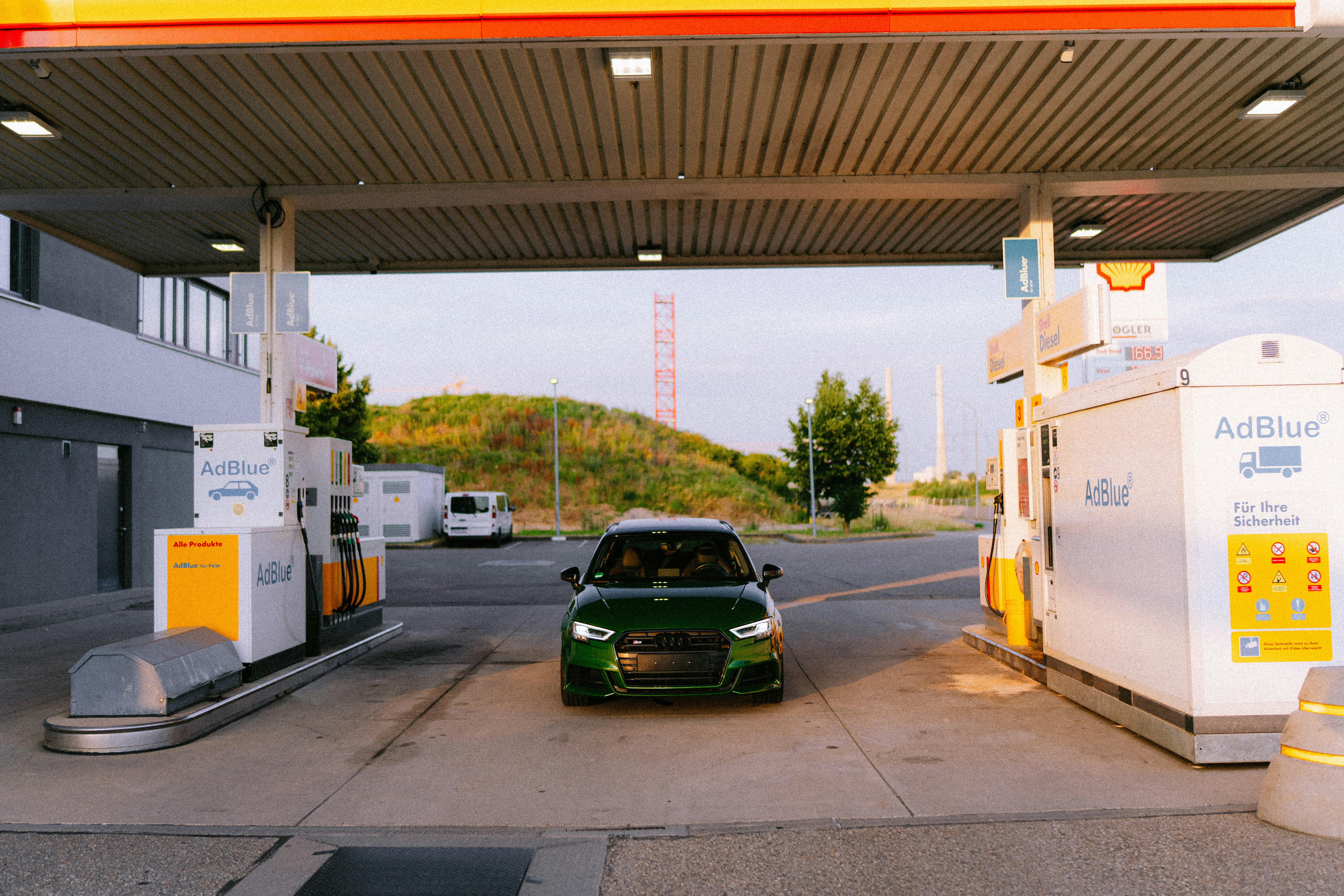 Green sedan at a sunlit gas station framed by canopies and distant hills