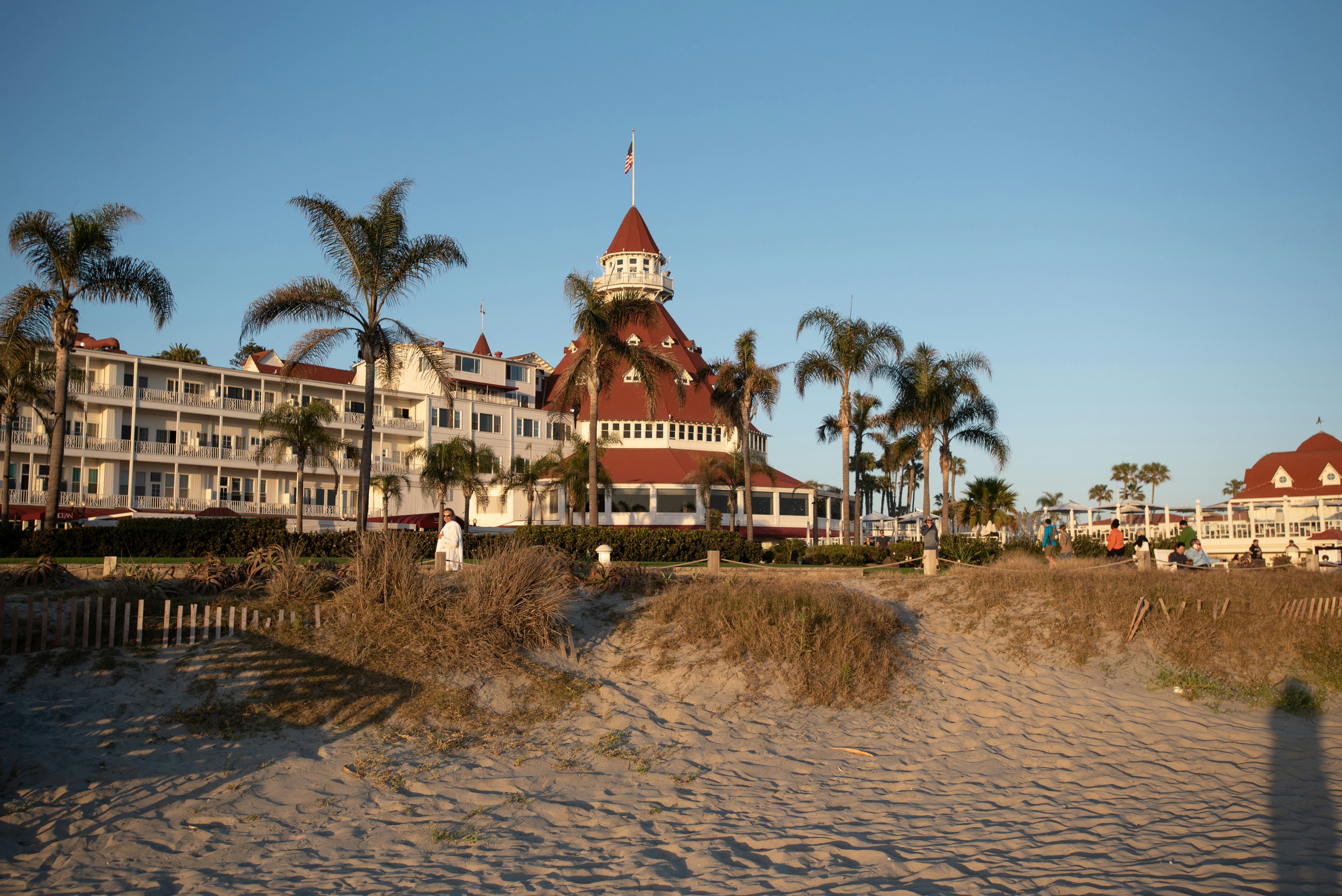 Sunlit beach scene with dunes in the foreground and a grand red-roofed hotel structure framed by palm trees along the shoreline.