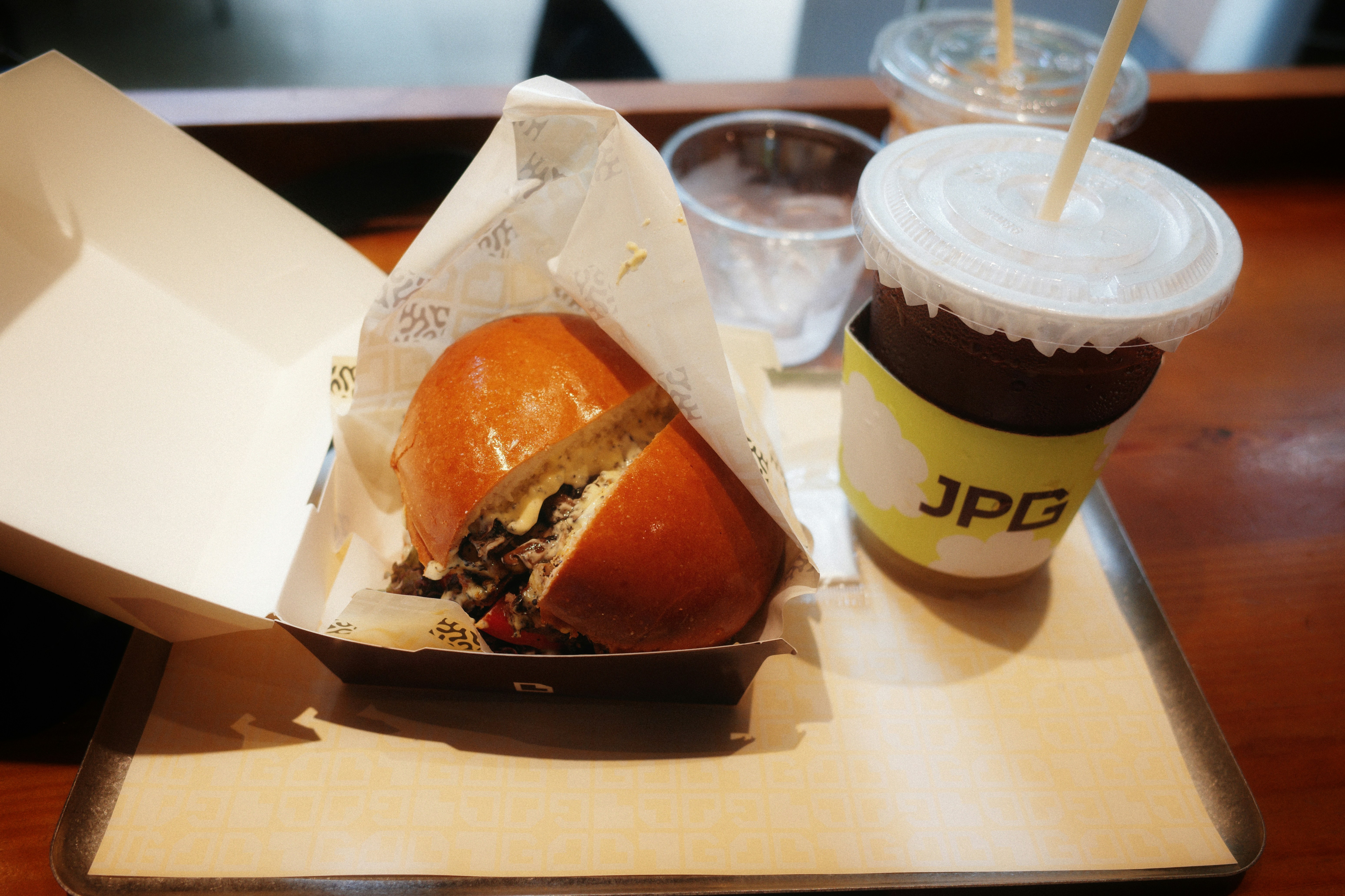 A fast-food burger wrapped in paper sits in a box on a metal tray beside a cold cup with a straw; a beverage cup with a logo is placed to the right. The scene is lit by natural light from a window, capturing a casual cafe meal setup.
