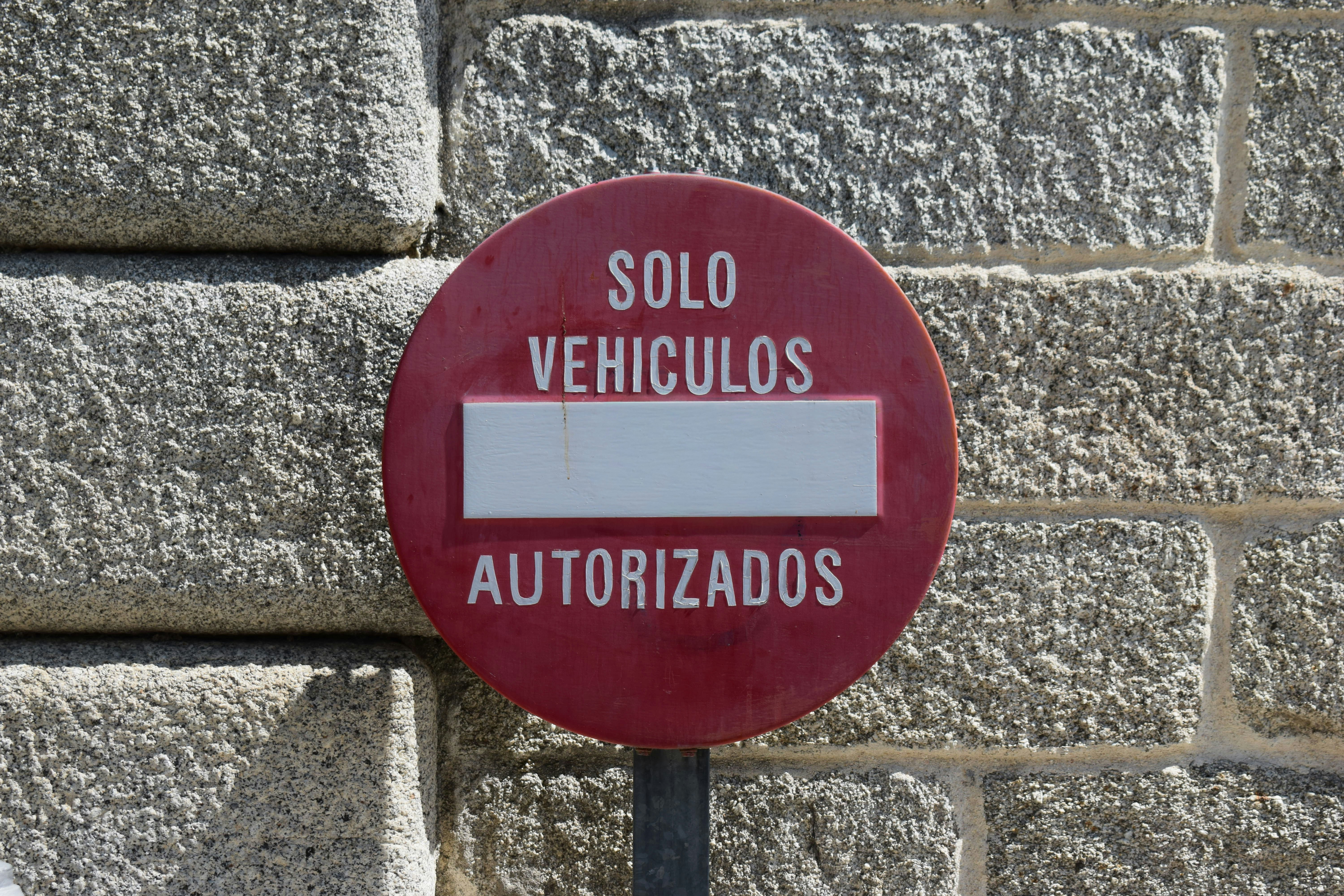 Red circle sign with white text and a blank panel mounted on a textured stone wall. Indicates that only authorized vehicles may enter; the sign is centered in frame with sunlight revealing wall texture.