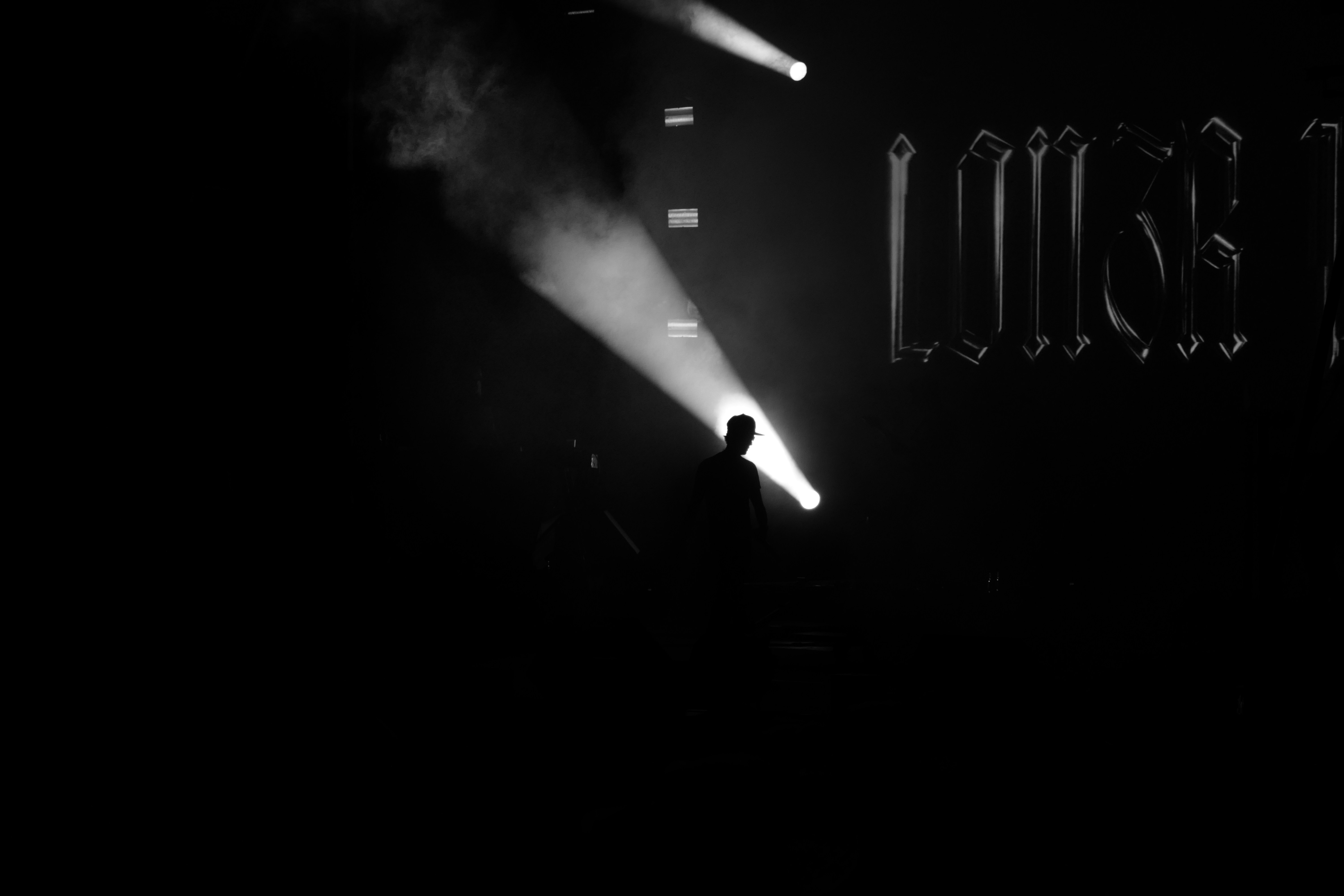 Dark concert scene with a lone silhouette illuminated by a strong stage beam; stage backdrop hints at band name. This is a photographic capture of a performance, with emphasis on atmosphere and light play.