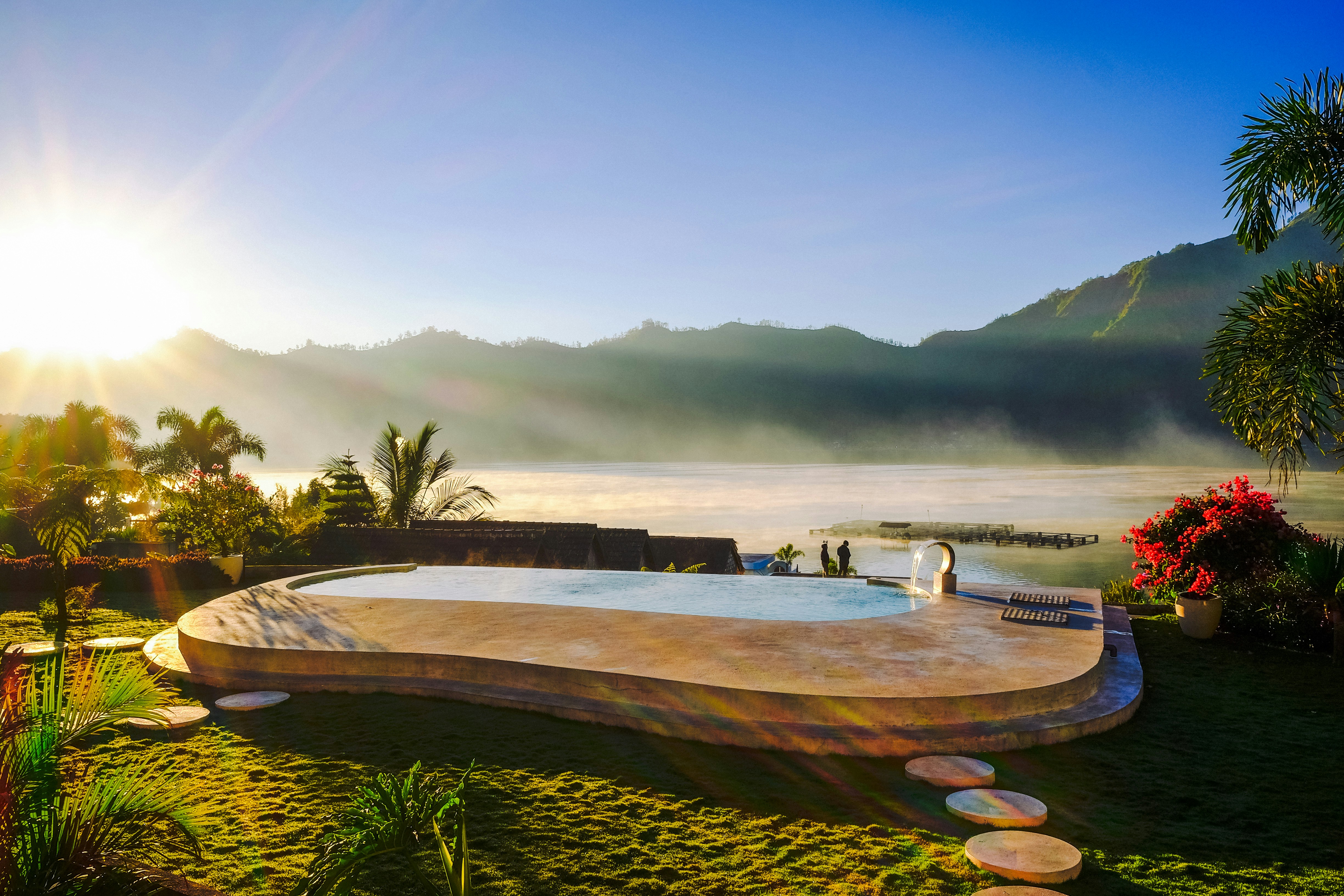 Sunlit infinity pool and curved terrace overlook a misty landscape with distant hills and a bright morning sky.