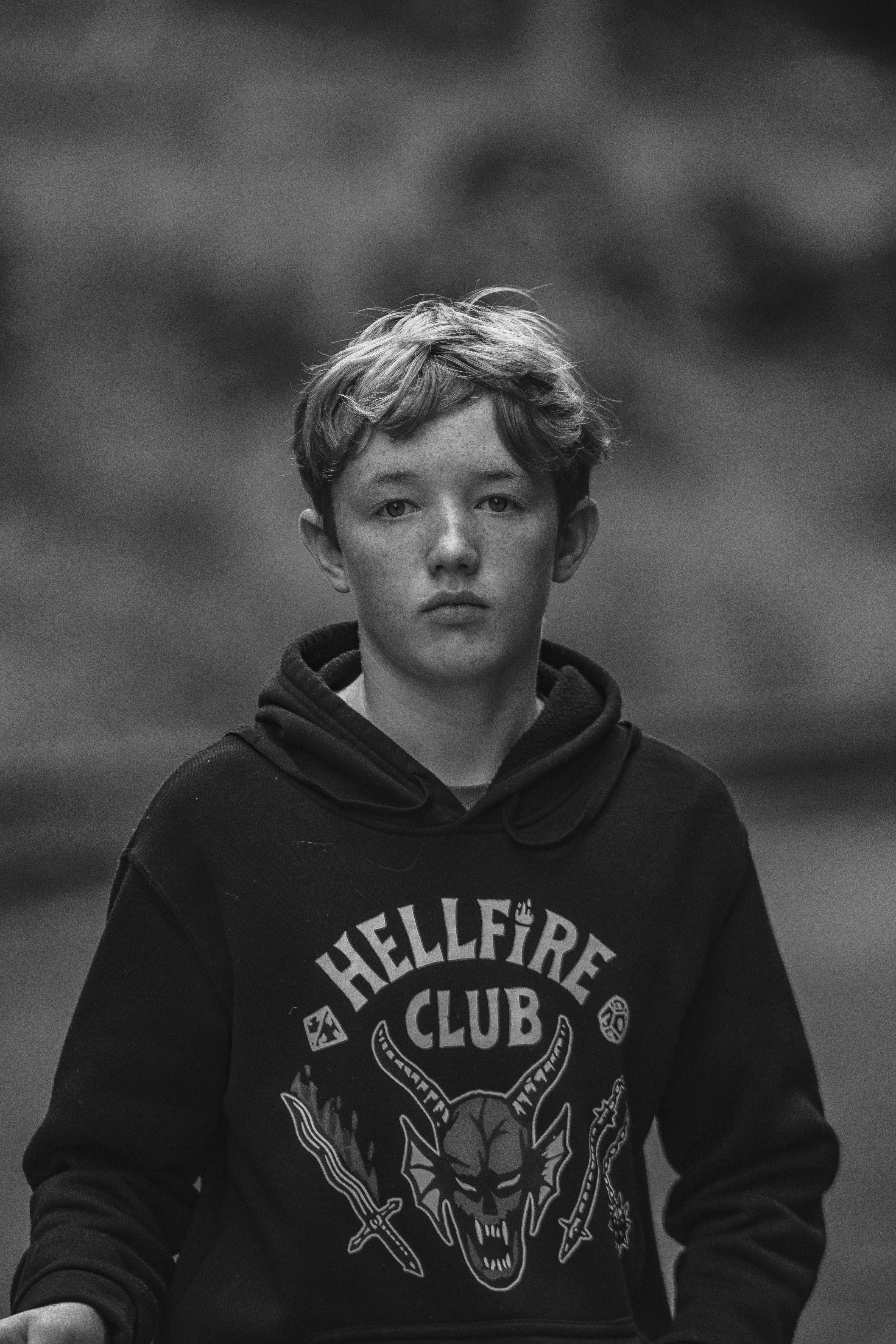 Monochrome portrait of a teenage boy wearing a hoodie with a Hellfire Club design, standing outdoors with a soft, blurred background.