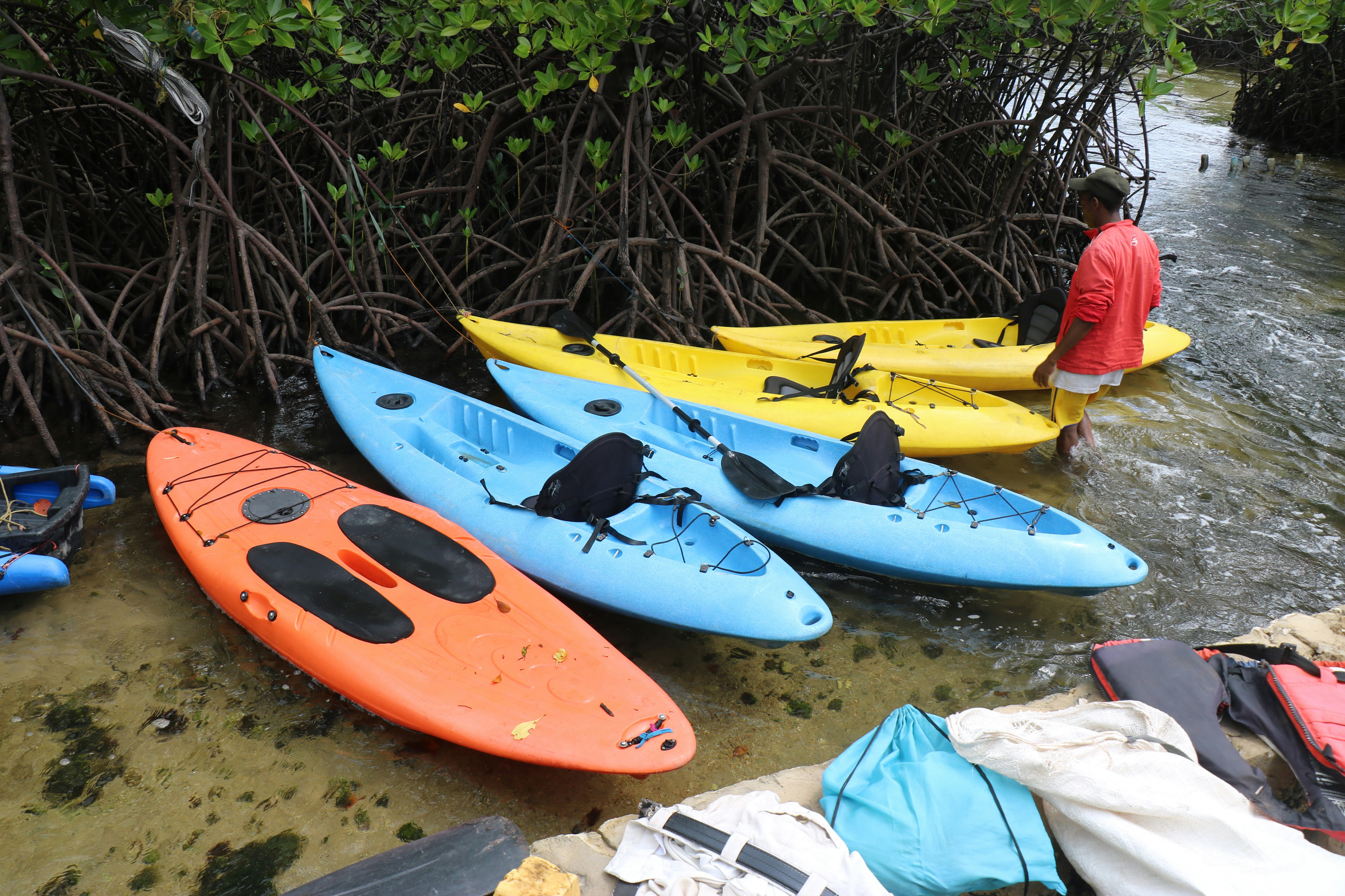 Several colorful kayaks resting on a shallow shoreline with a person in a red shirt standing near the water. The scene suggests a dock or launch point with gear laid out nearby.