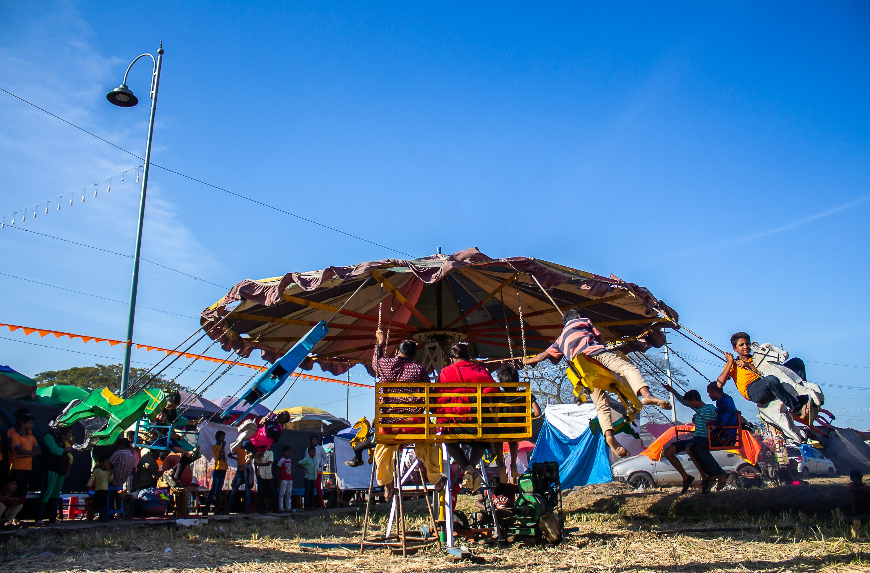 Vibrant carnival scene with a circular ride canopy and people gathered around in summer light. A lively mix of tents, riders, and spectators create a dynamic, colorful snapshot.