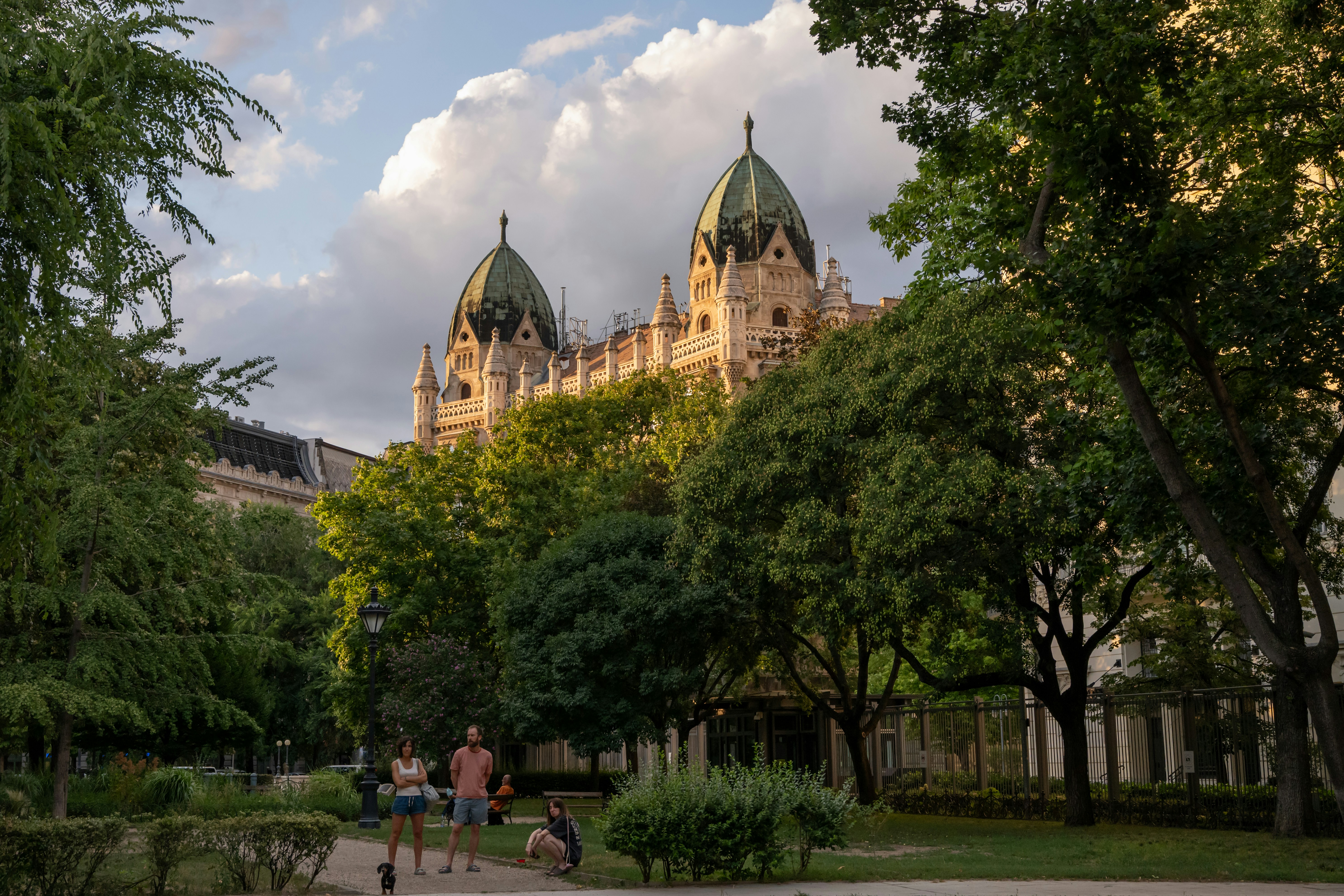 Sunlit domed towers rise above a tree-lined park with visitors in the foreground, capturing a calm urban moment near a historic building. (photography print)