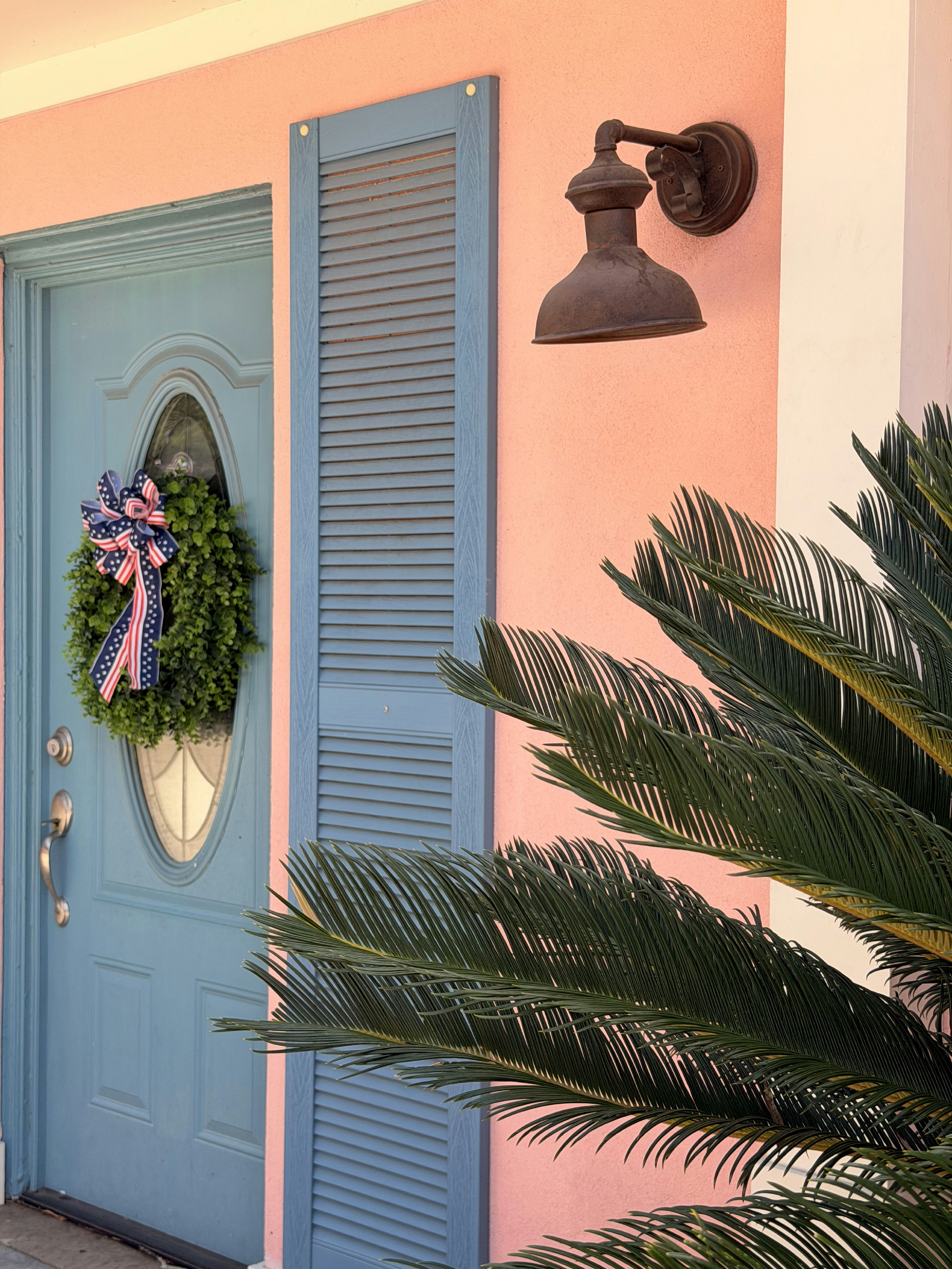 Pastel pink wall with a blue front door and shutters, accented by a wreath and a rusted exterior lamp, framed by tropical plant fronds.