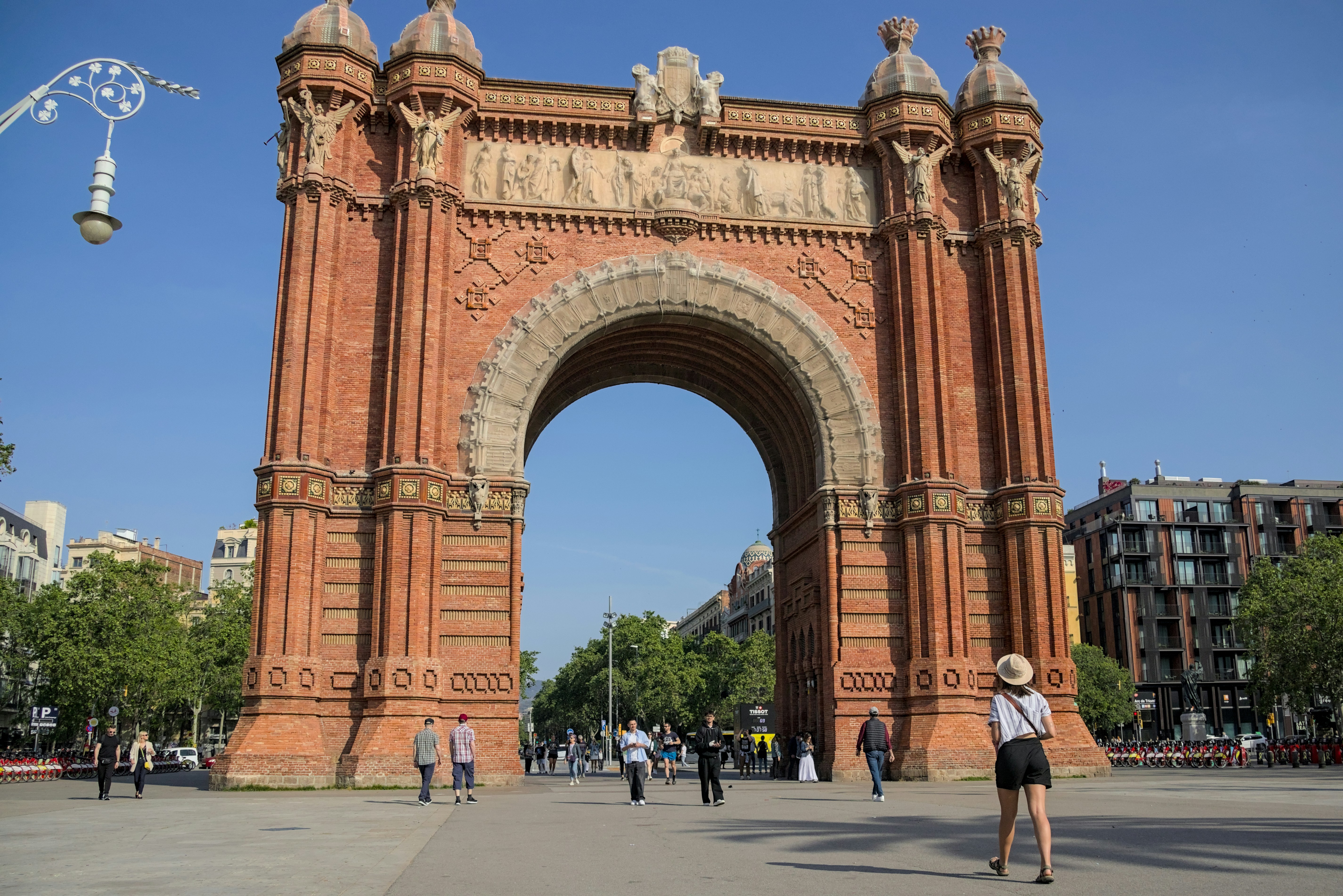 Red brick triumphal arch with people strolling in a plaza, city buildings in the background under a clear blue sky.