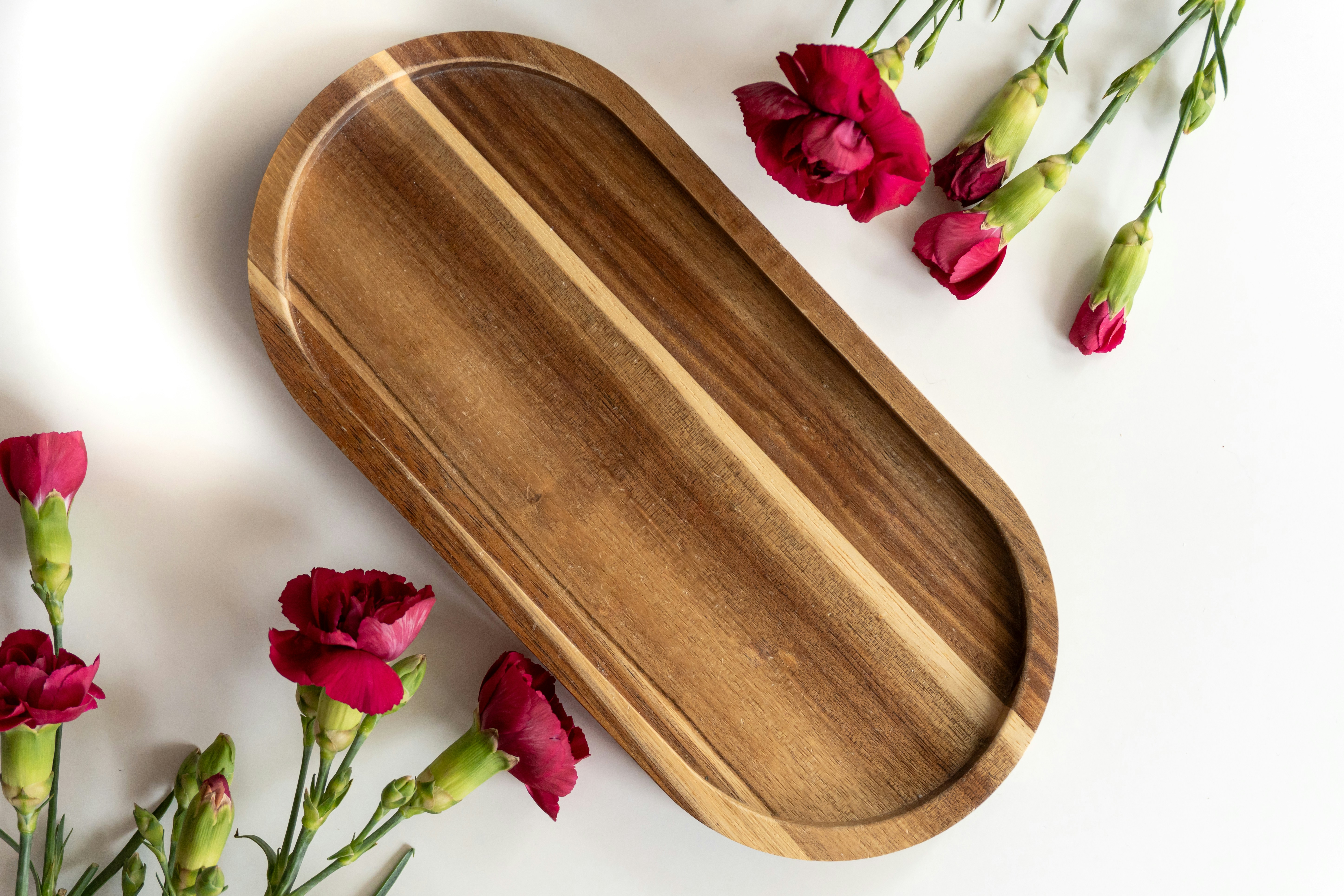 Wooden oval tray on a light background, surrounded by pink carnations; a still-life composition highlighting texture and color contrast.