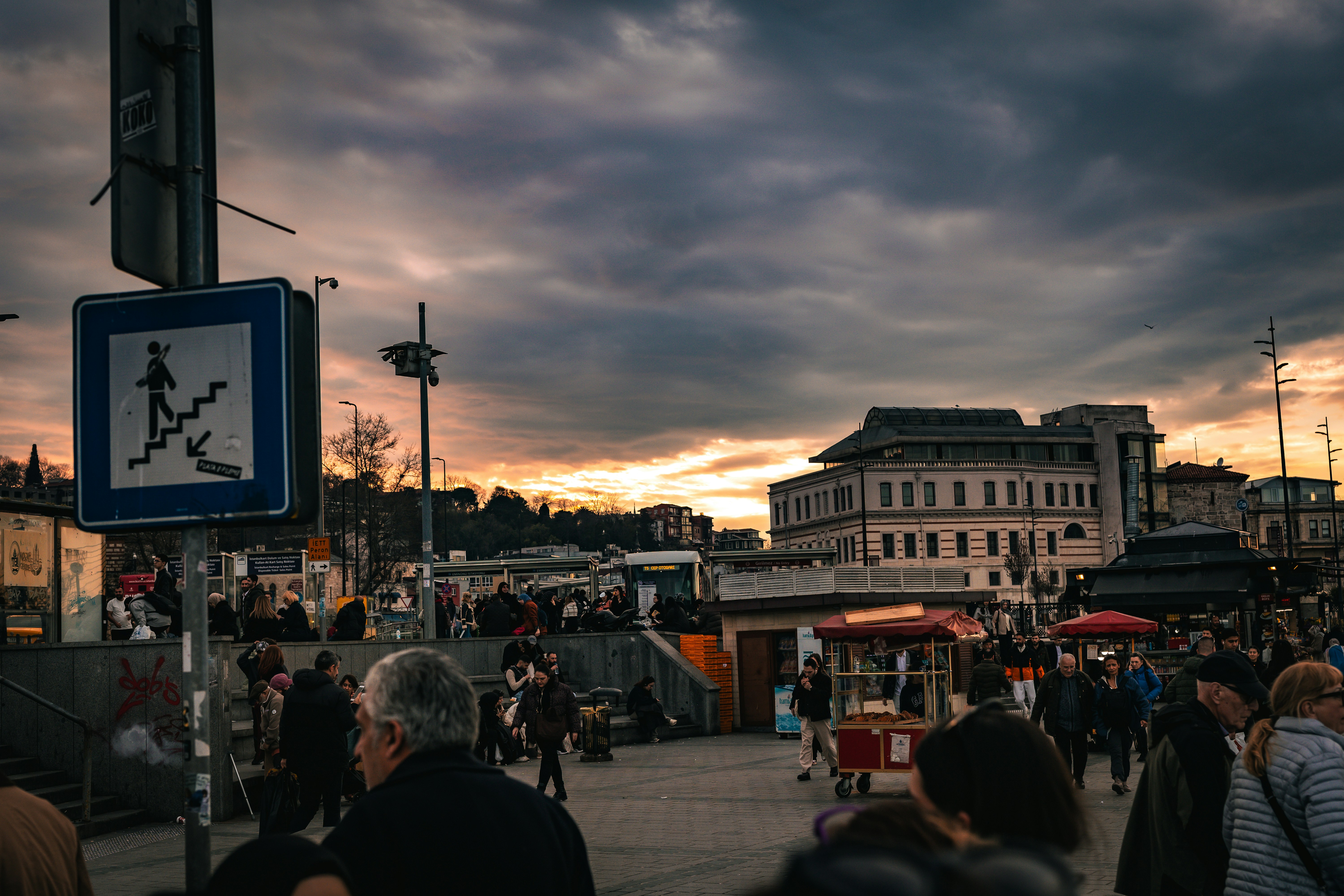 Bustling urban square at dusk with people milling about, market stalls, and a dramatic sky over classic buildings.