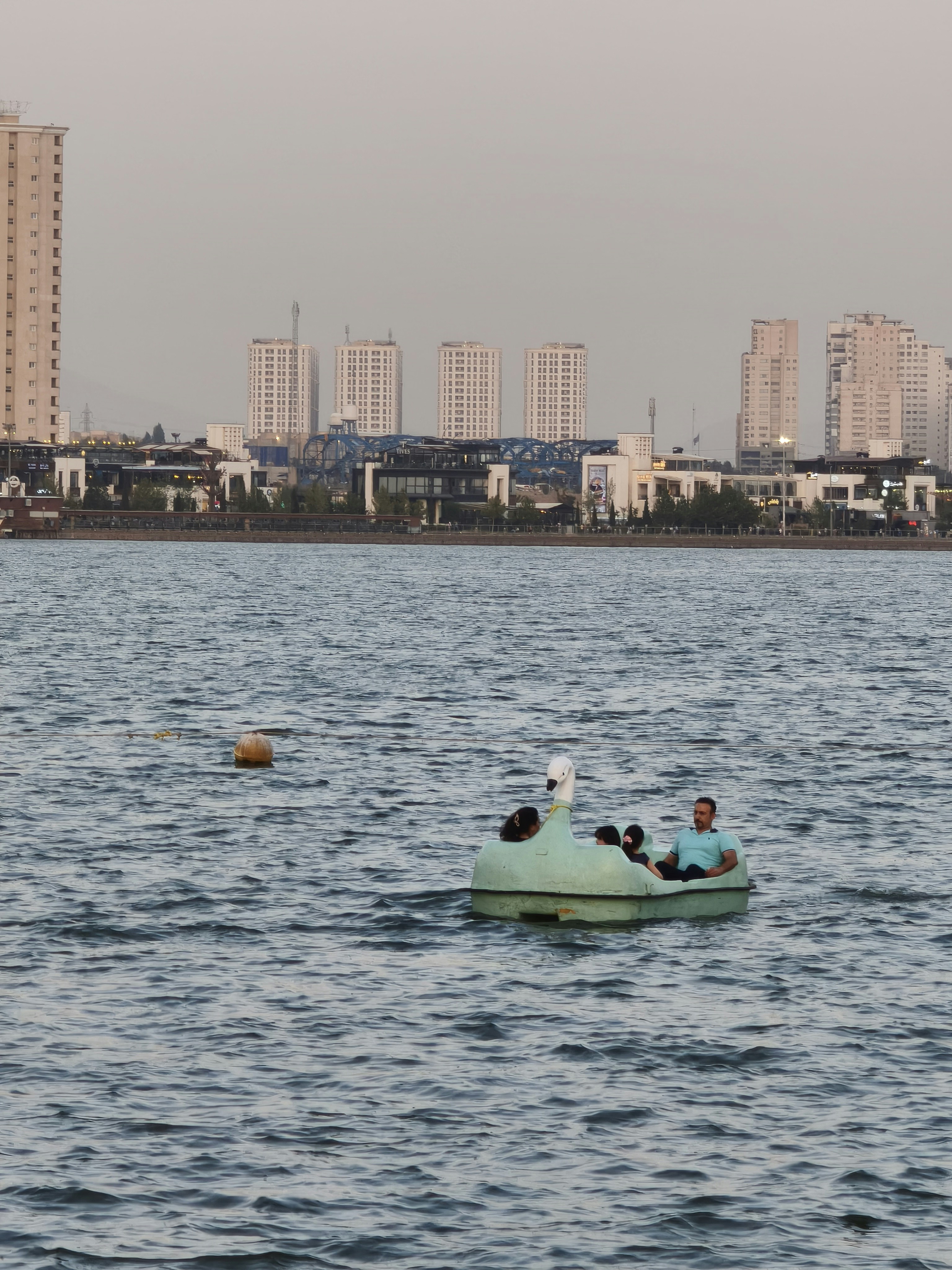 A small green paddle boat carries four passengers across a calm coastal water with a city skyline in the background.
