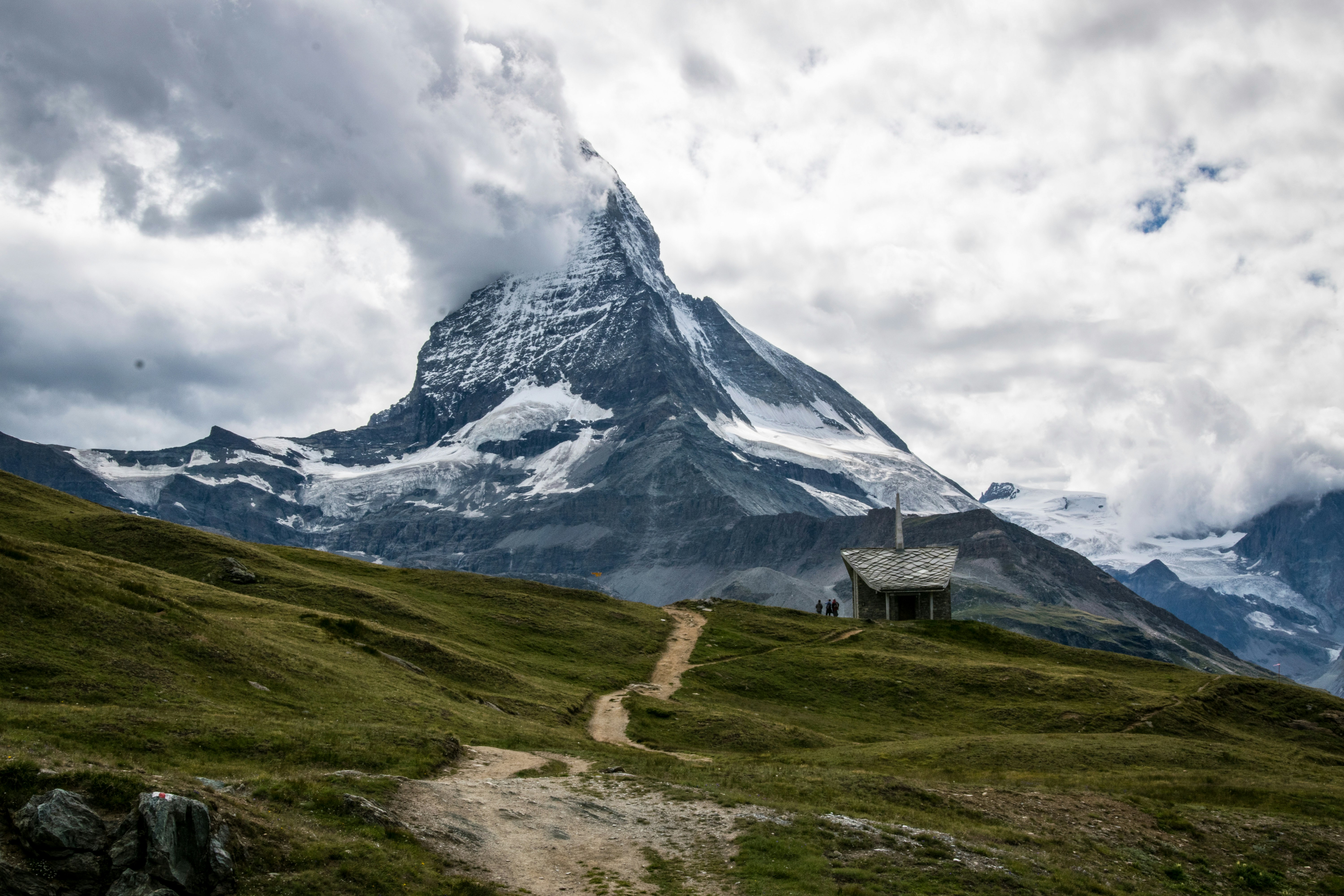 An alpine path leads to a small cabin with a dramatic mountain backdrop and dramatic clouds.