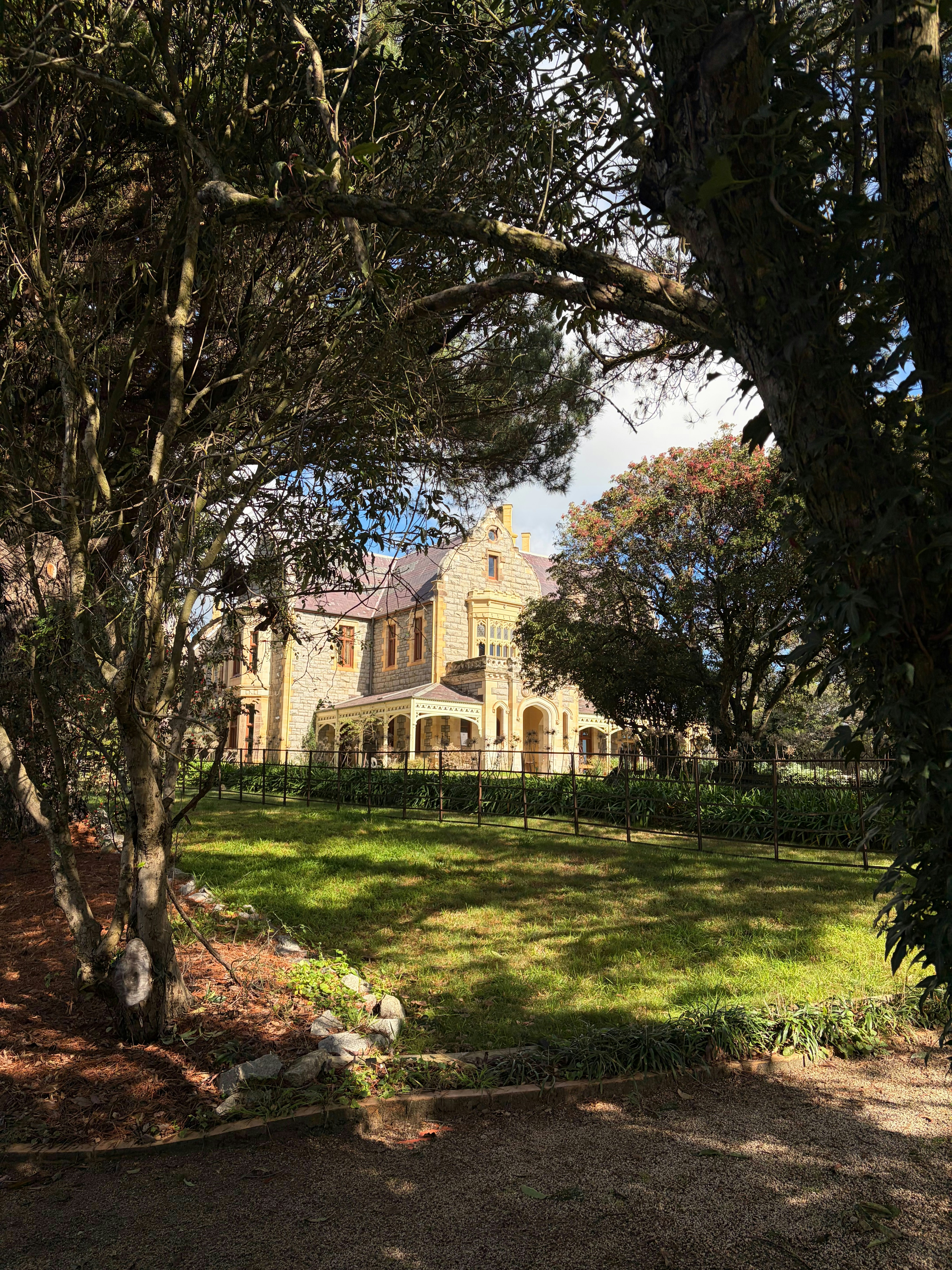 Sunlit villa with ornate stone facade framed by trees and a manicured lawn, visible through a park-like garden.