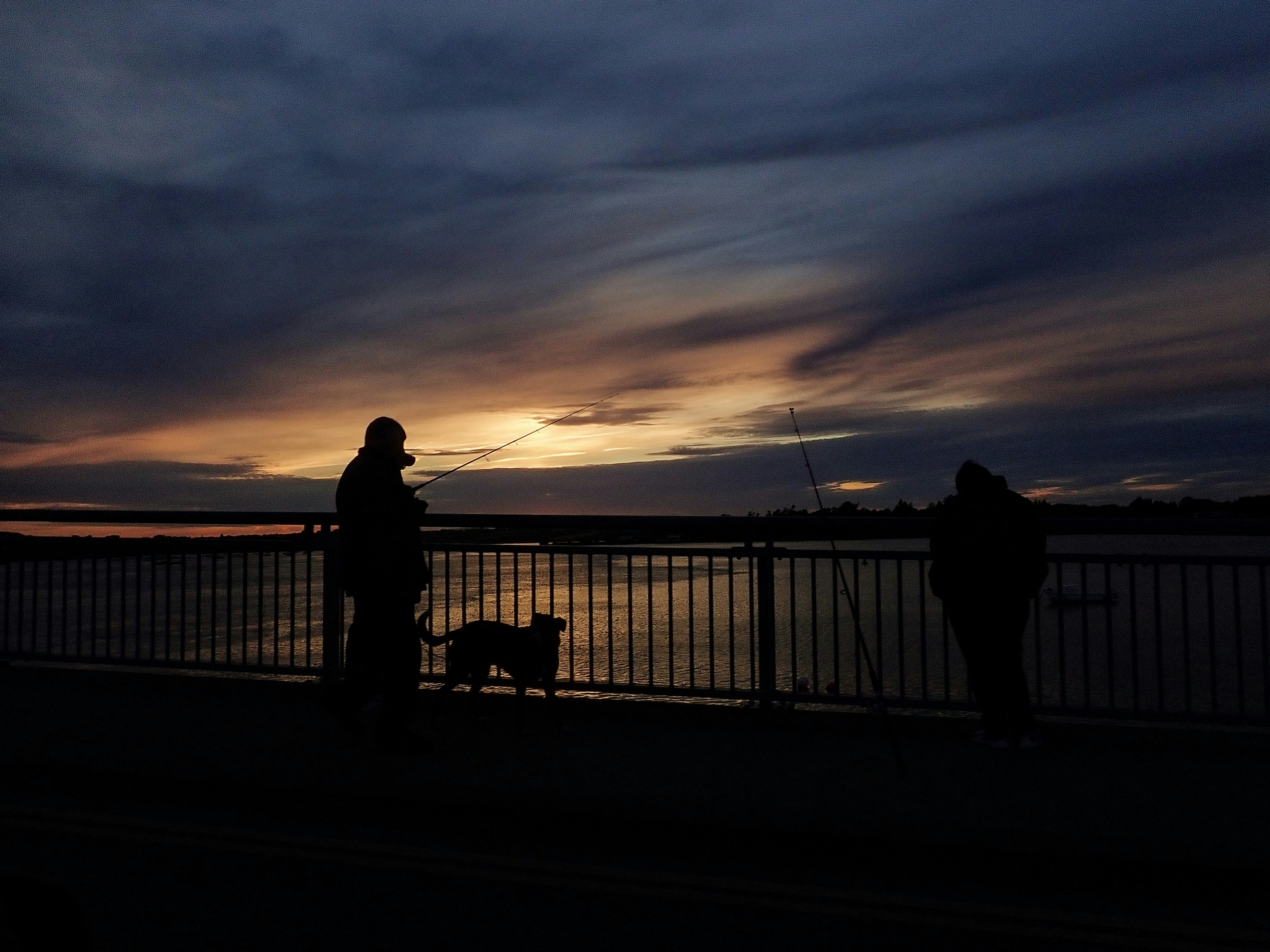 A peaceful evening scene on Wexford Bridge, Ireland, as twilight settles over the water. The striking silhouettes of two fishermen and their dog are framed perfectly against a dramatic sunset sky, where deep blue clouds split to reveal a vibrant streak of golden light. A calm, atmospheric moment capturing the timeless rhythm of local life and outdoor leisure at dusk.