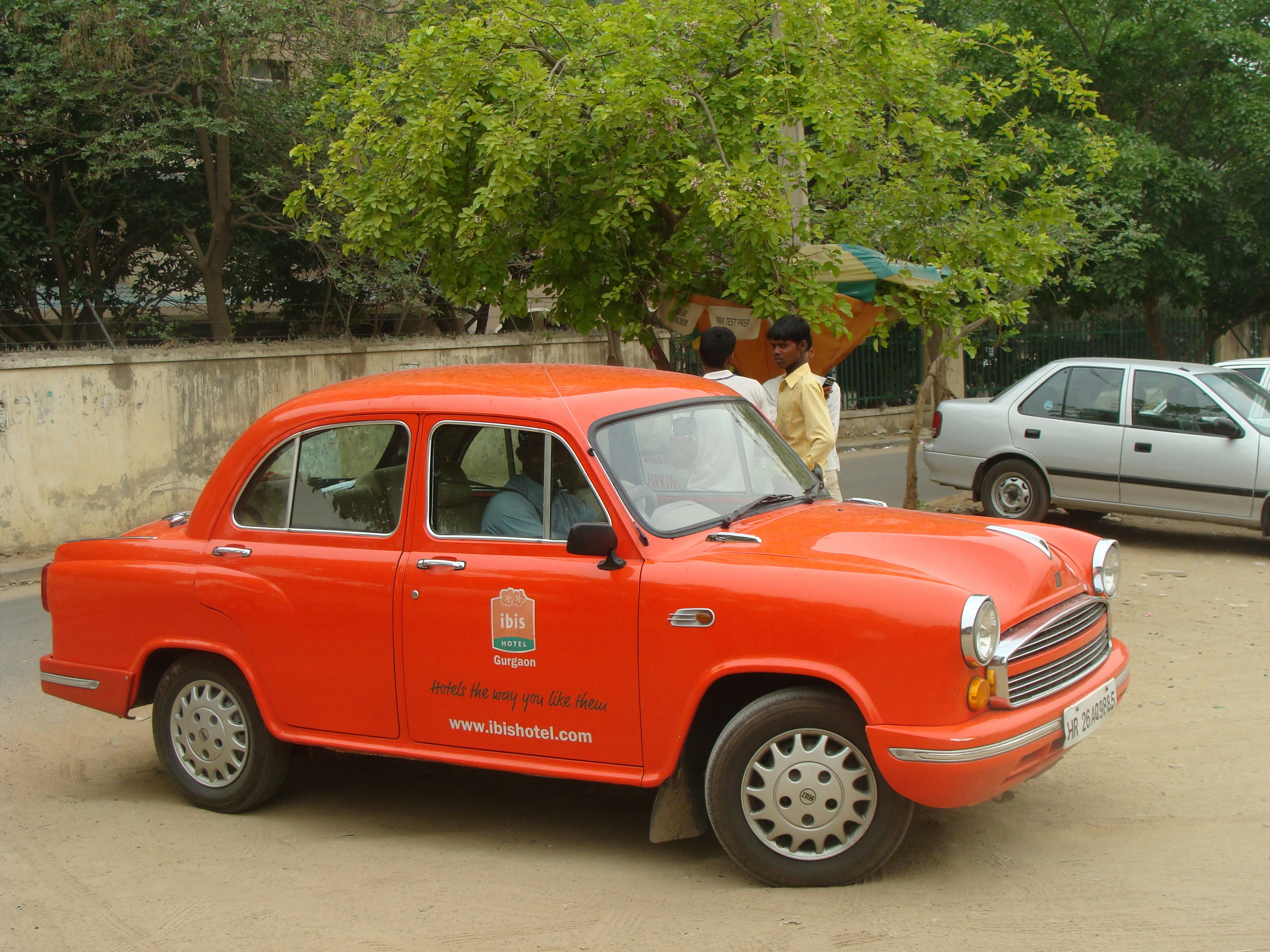 Orange vintage sedan parked on a dusty street with pedestrians in the background