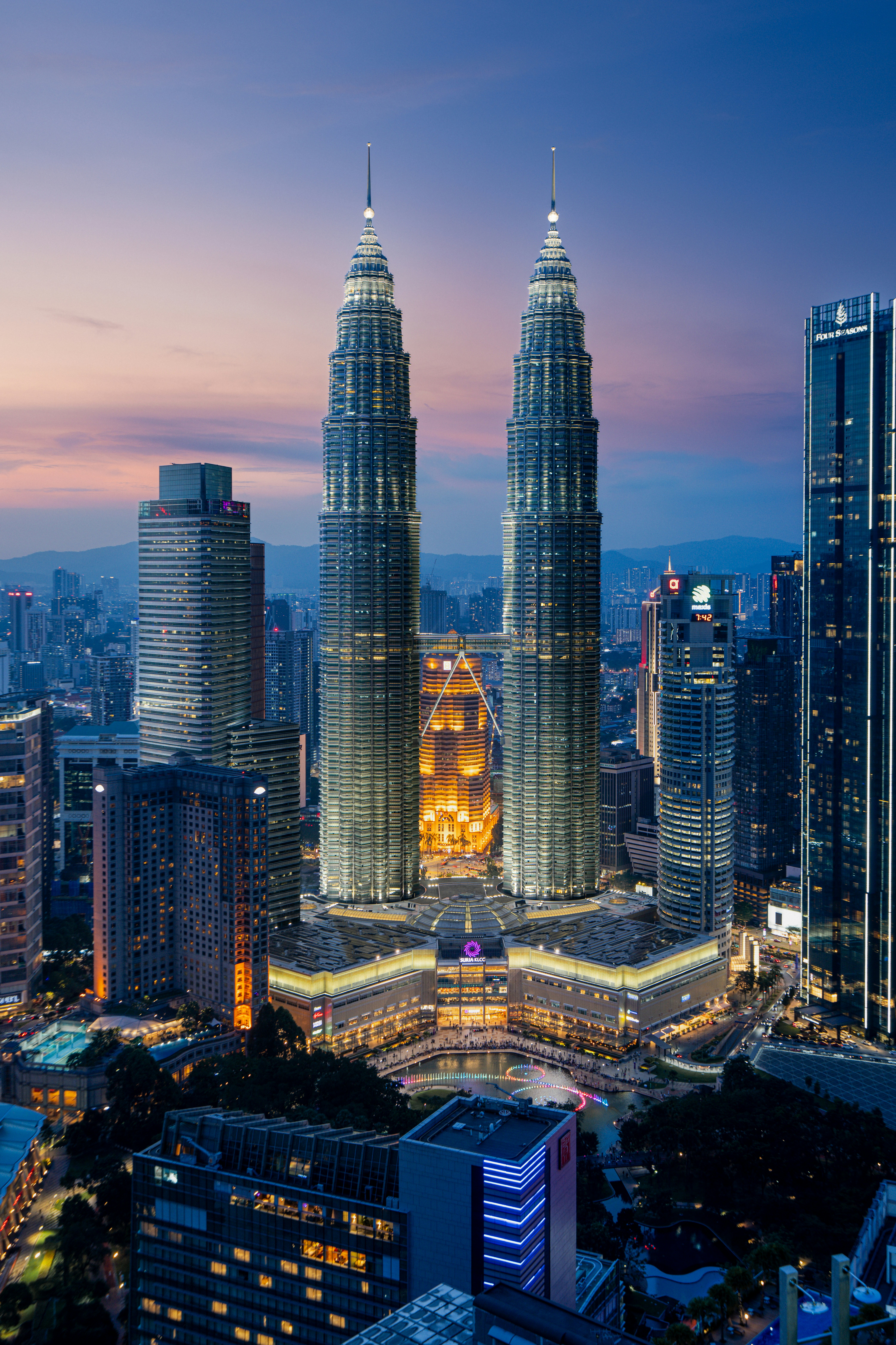 Twilight view of the Petronas Twin Towers rising above the city, illuminated against a gradient sky with surrounding skyscrapers and a landmark-lit plaza below.