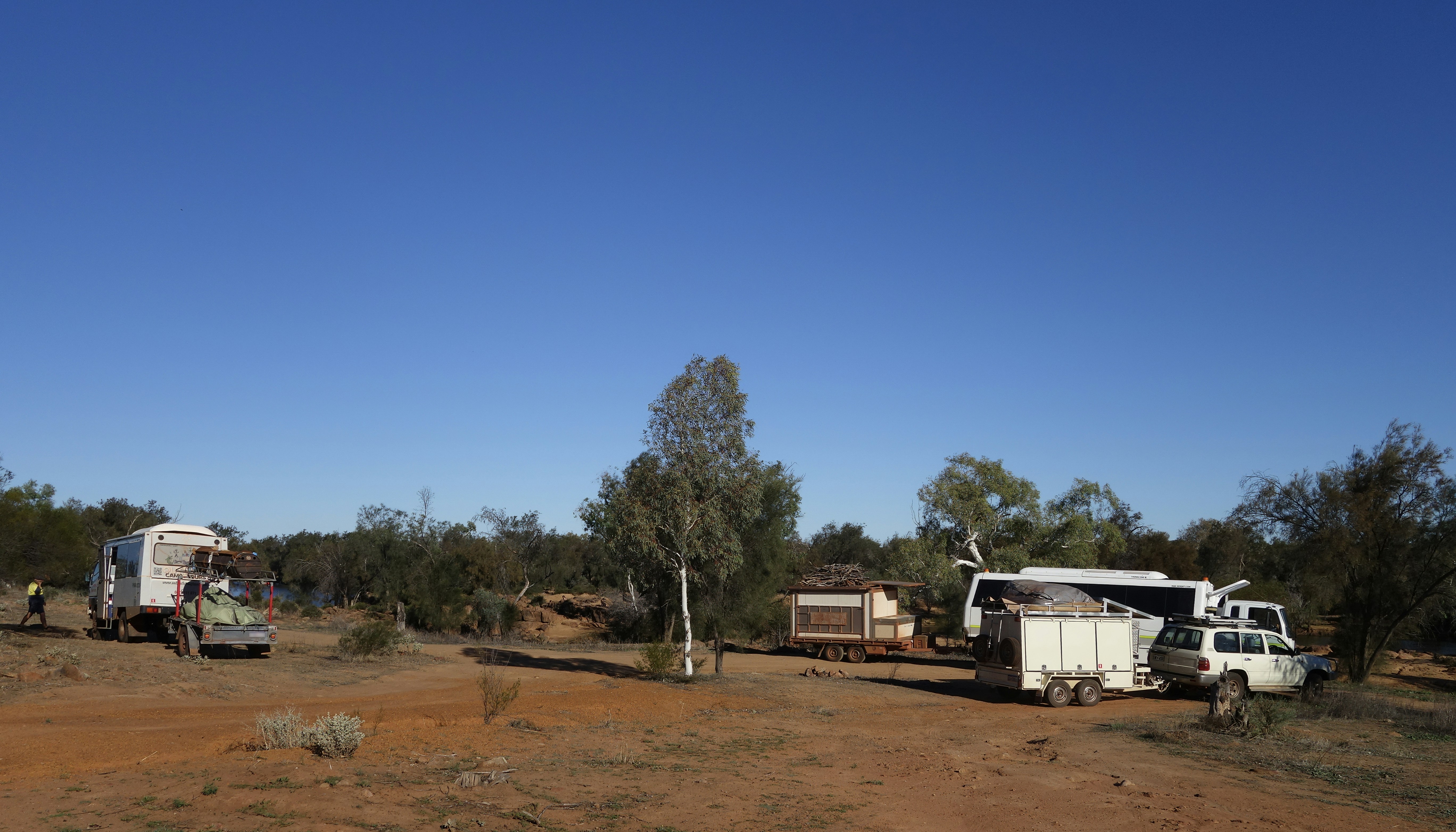 Rugged caravan park with campers and trucks set on red desert soil under a wide blue sky. A small cluster of trees and scattered vehicles anchor a remote, sunlit encampment.