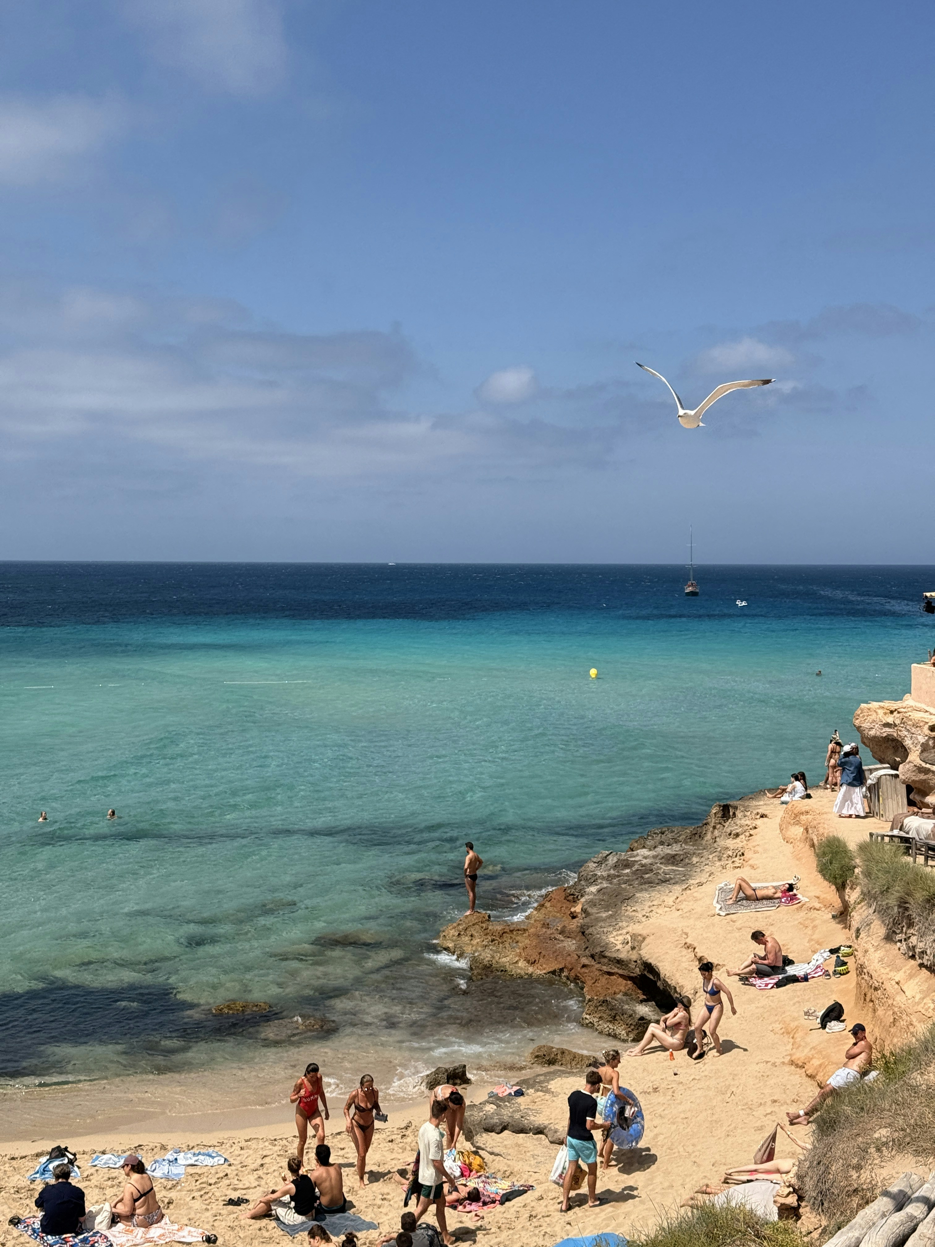 A sunlit beach scene with turquoise water, rocky shoreline, and swimmers enjoying the sea under a clear sky.