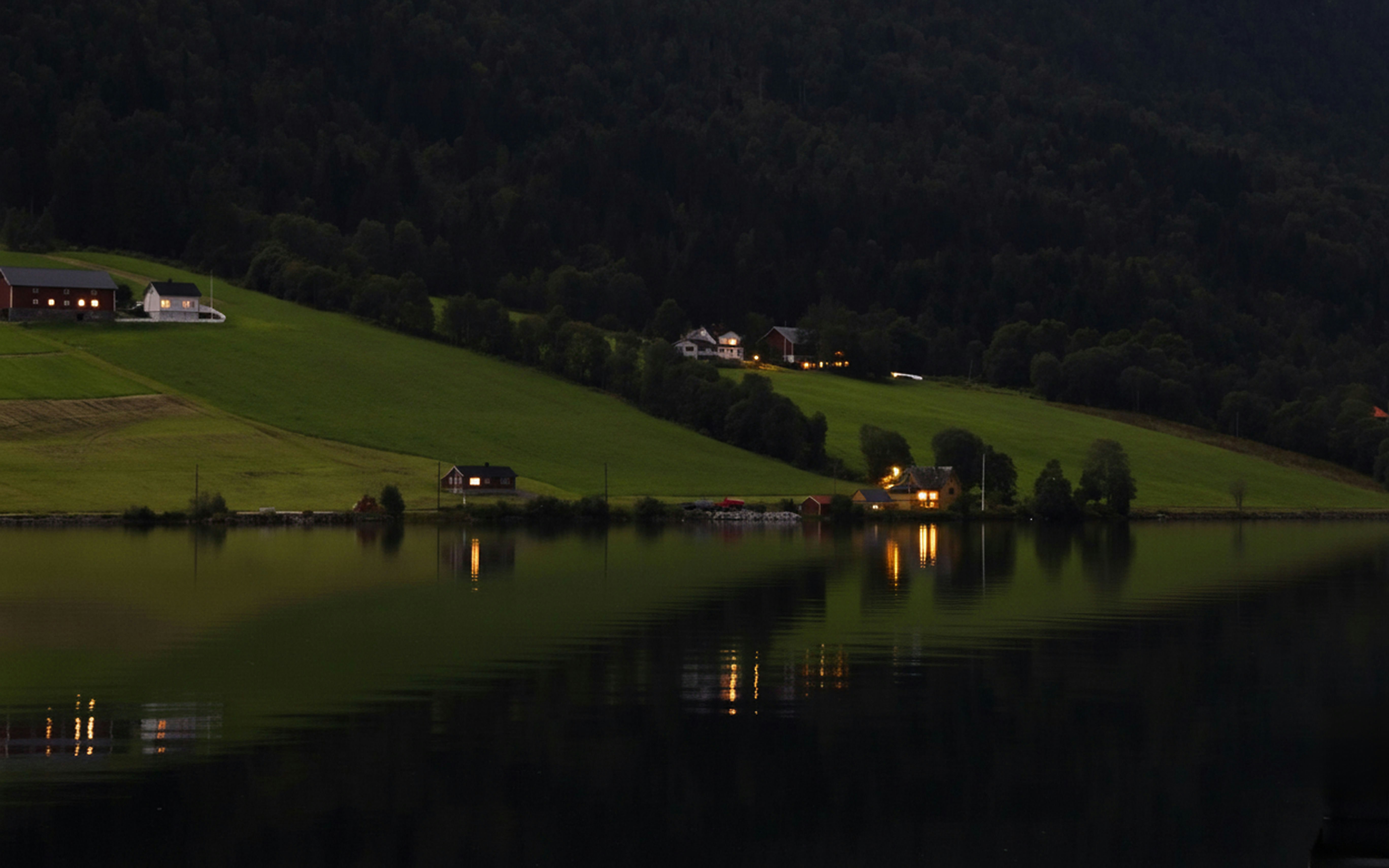A tranquil evening landscape along a Norwegian fjord, where scattered countryside homes glow warmly against lush green hills and a dense forest. Their reflections shimmer on the calm water, creating a peaceful and atmospheric scene that captures the beauty of rural Norway at dusk. Ideal for travel, nature, landscape, and Scandinavian-themed projects.