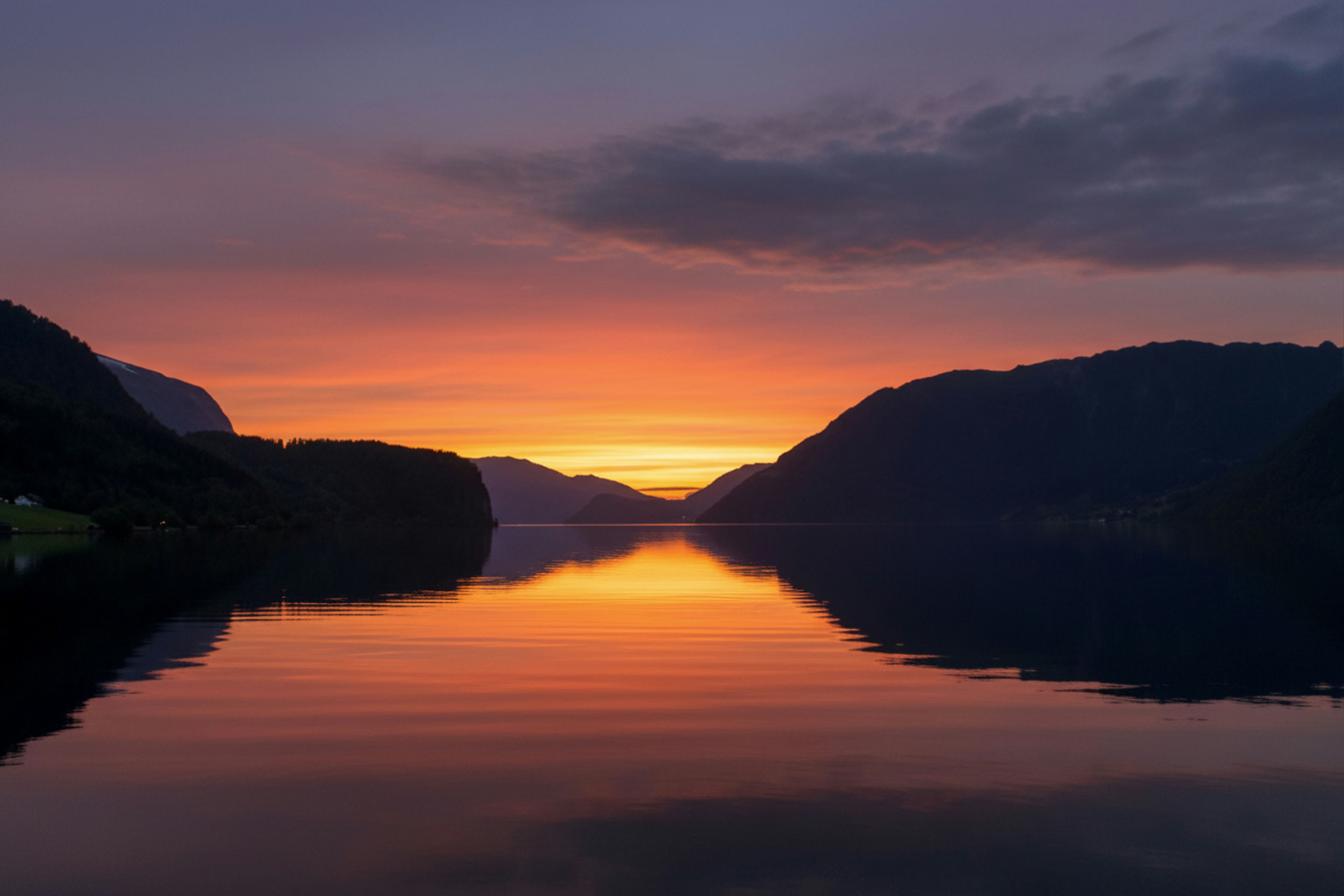 Vibrant sunset over a calm fjord with mirrored water and silhouetted mountains on both sides. A serene landscape emphasizing warm hues and reflections.
