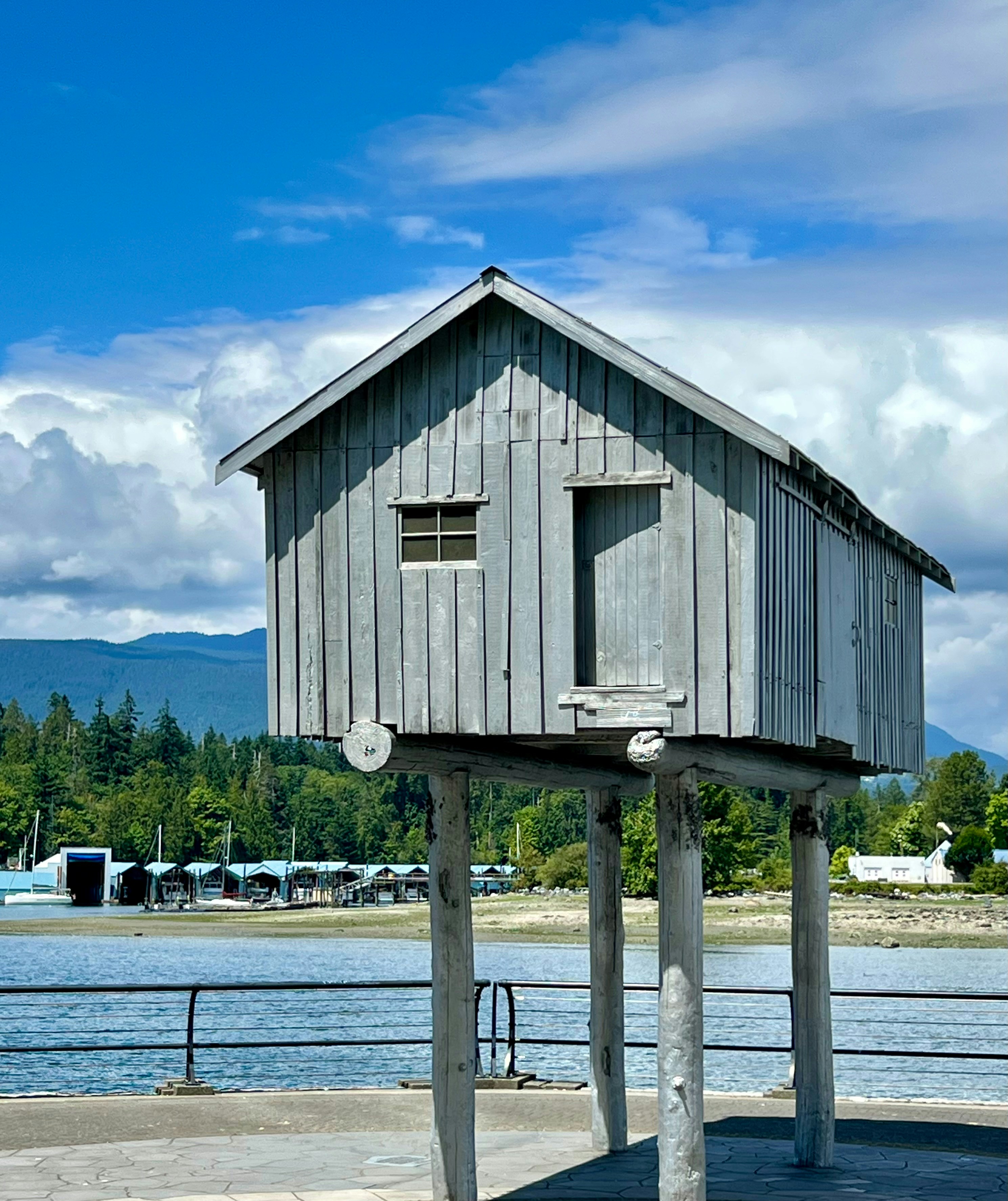 Weathered wooden shack on stilts stands over calm waterfront with distant hills and clear blue sky; a quiet coastal scene emphasizing rustic architecture.