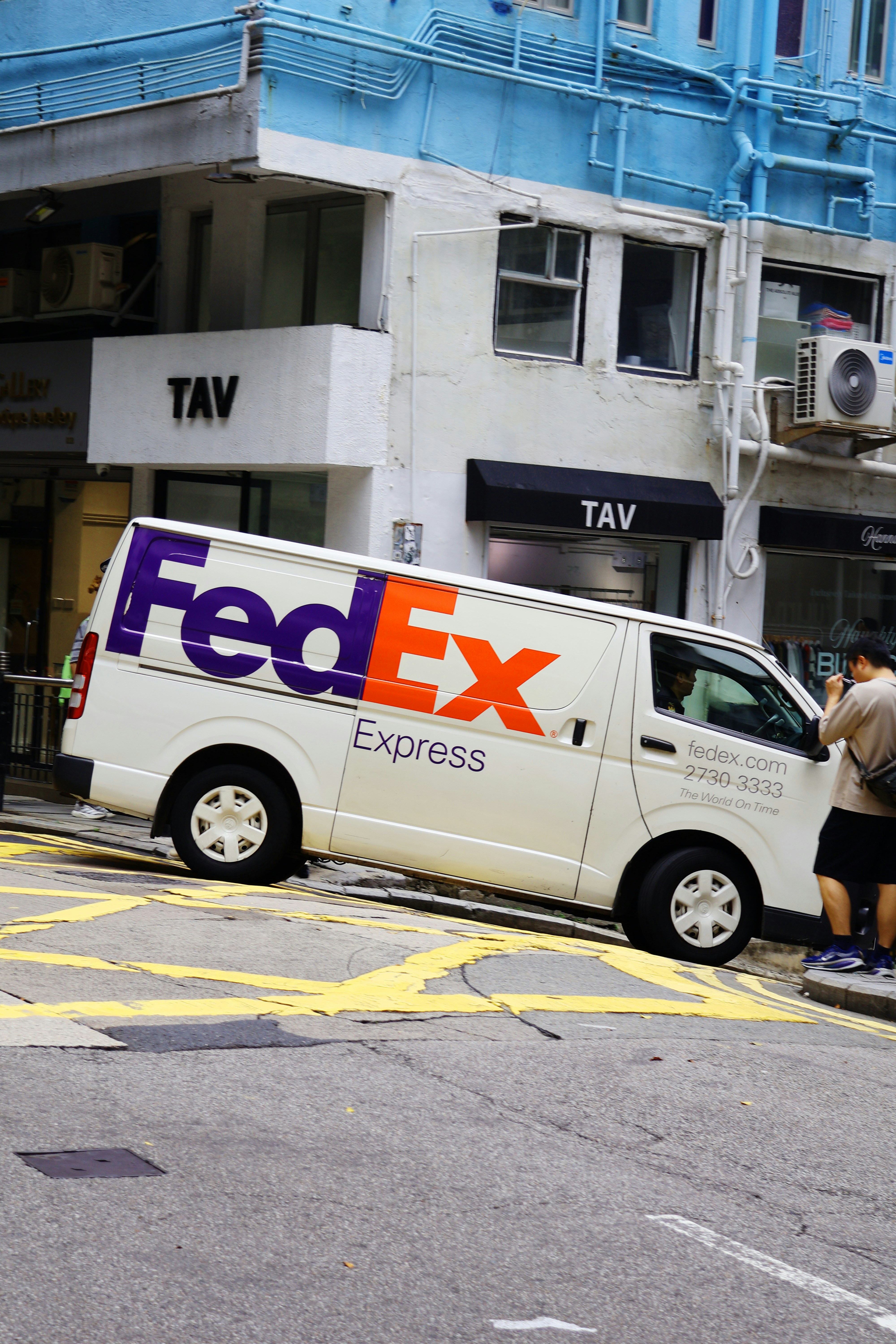 FedEx van in motion past a blue storefront on a slanted city street