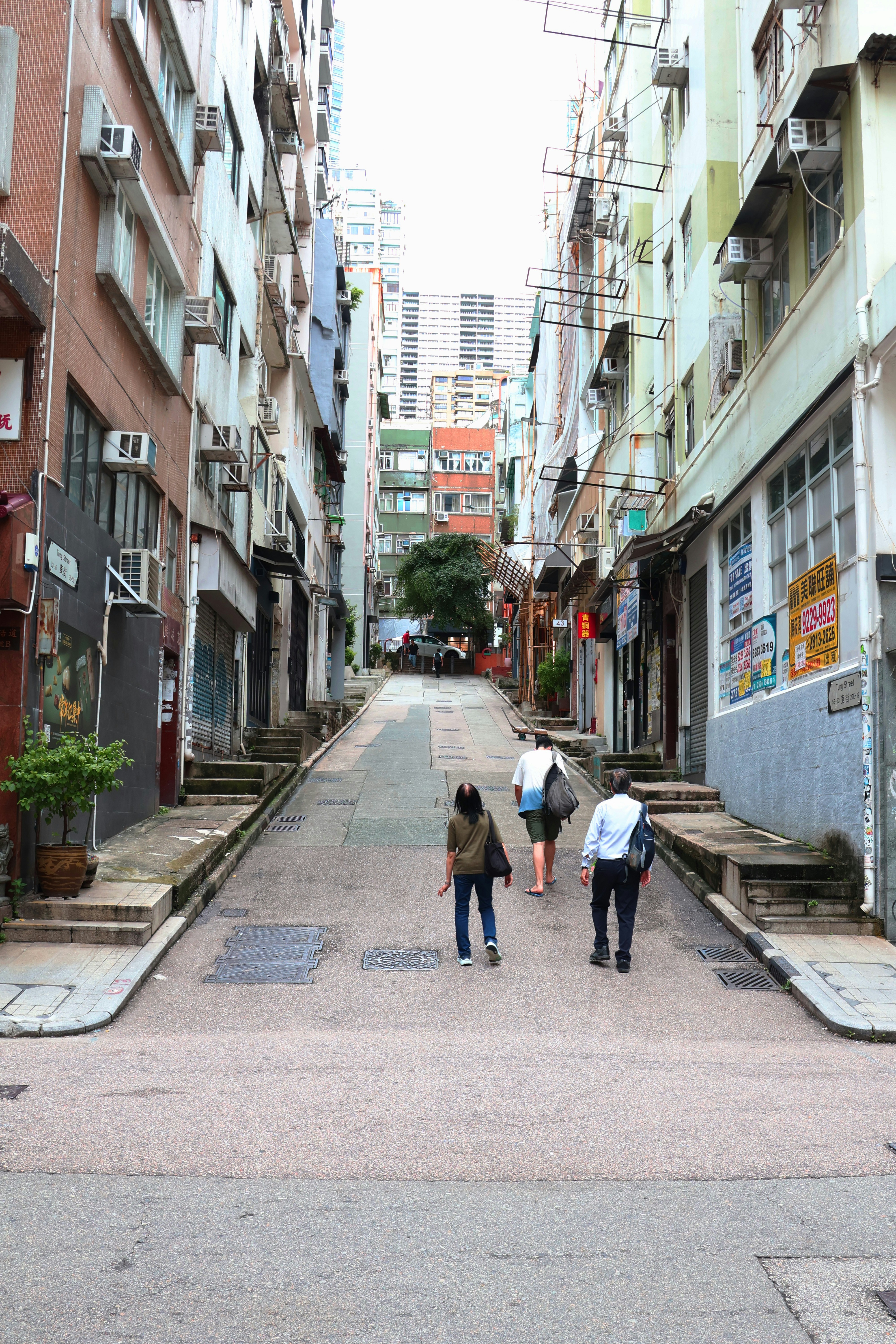 A pedestrian couple and a companion walk up a sunlit urban alley lined with colorful apartment buildings.