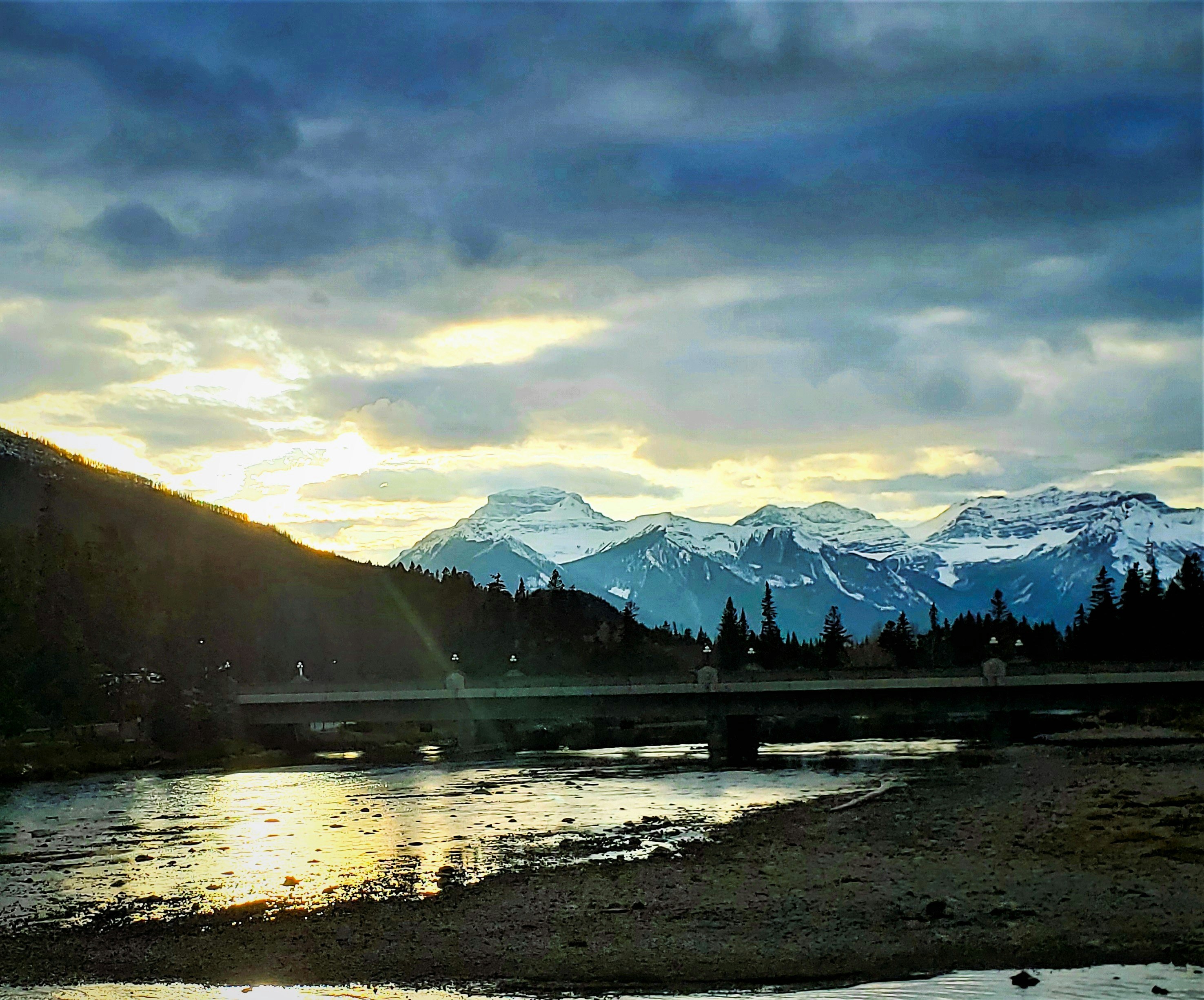 Snowy mountains rise behind a calm river and a bridge at dawn, with soft golden light reflecting on the water.