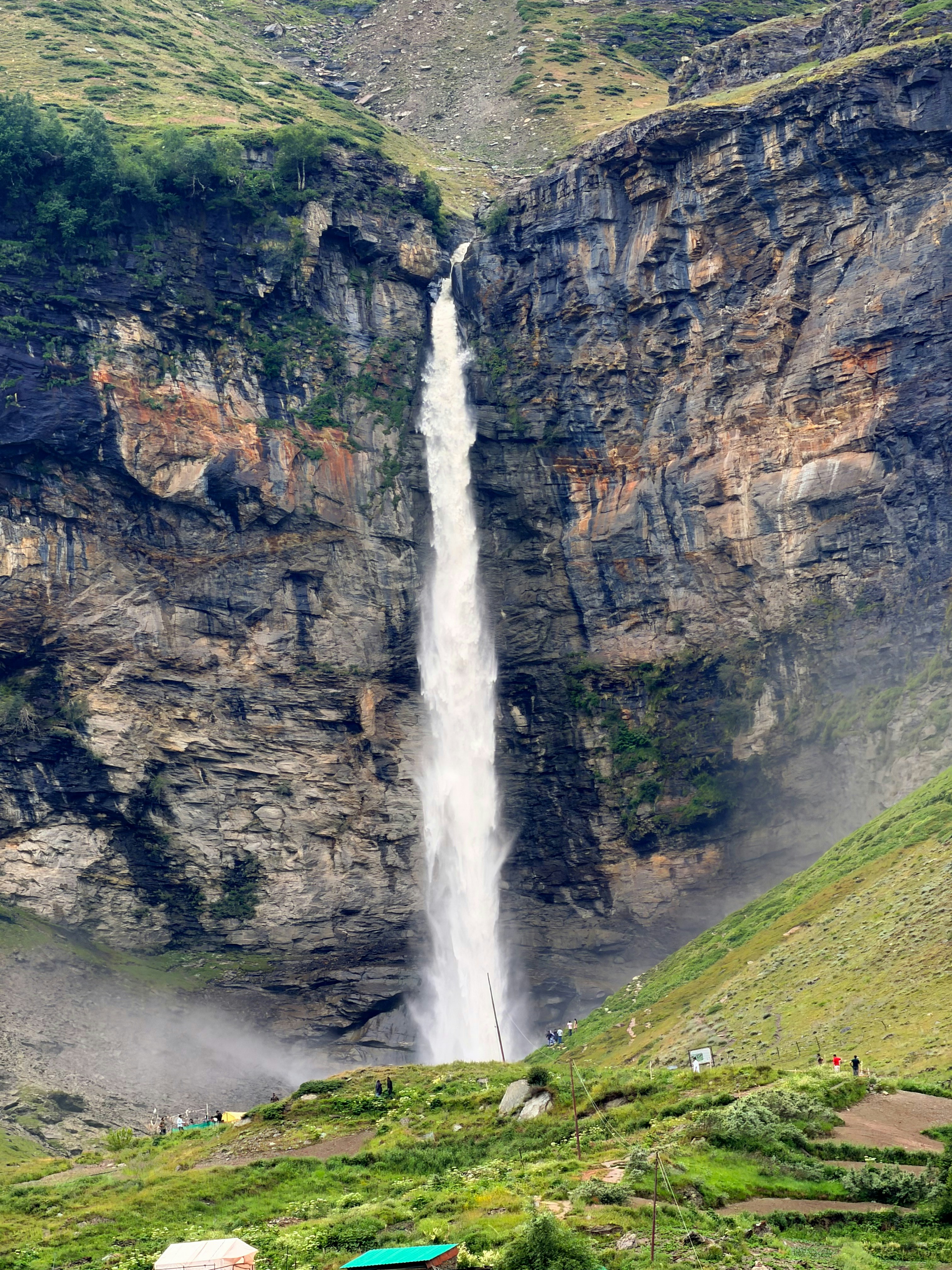 A tall, narrow waterfall plunges between rugged cliff faces, with mist rising at the base and a green valley dotted by small structures and pathways below.