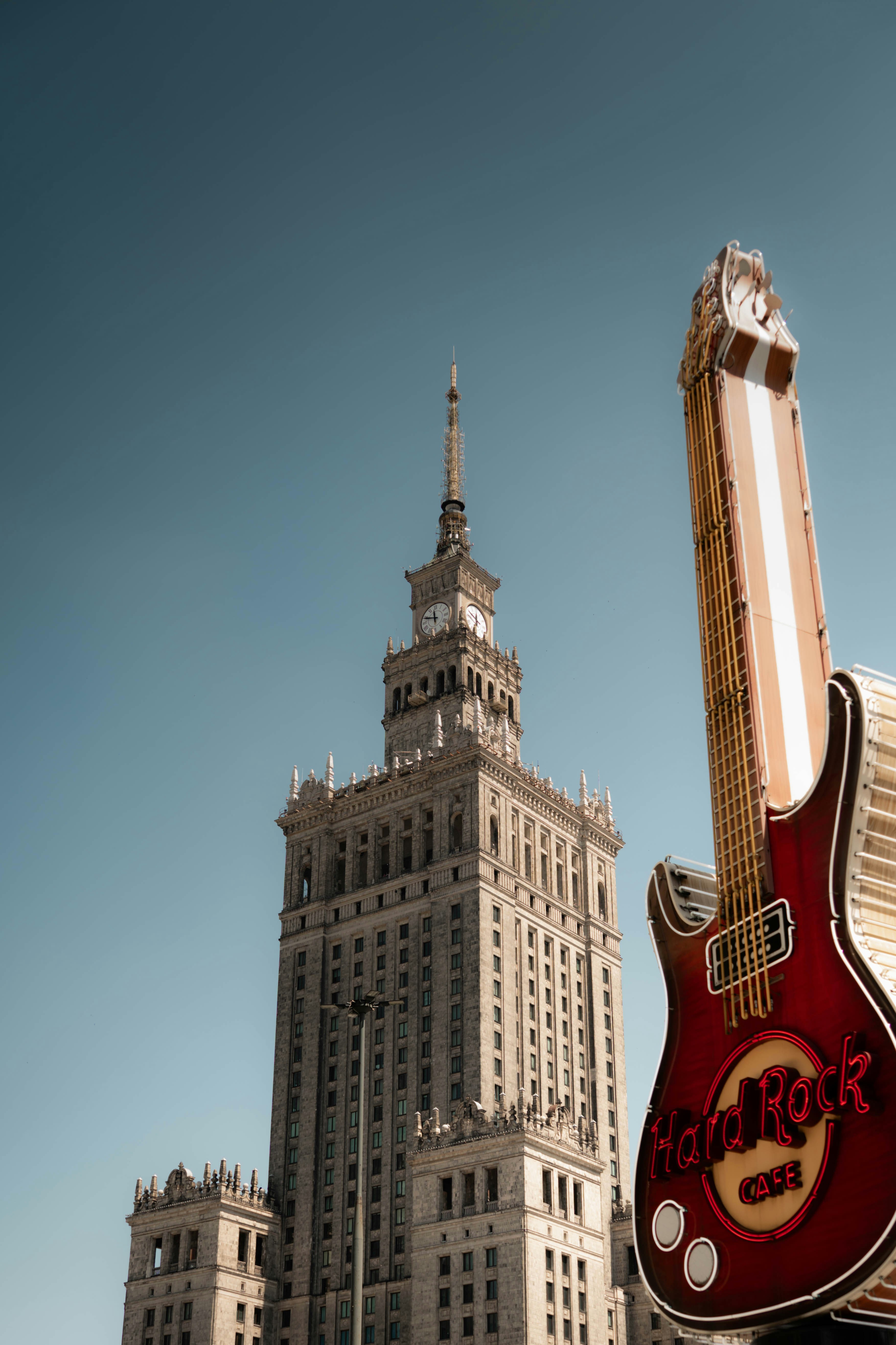 A city street scene featuring a towering historic building beside a large illuminated guitar sign, blending architectural grandeur with vintage rock iconography. The image captures contrast between timeless architecture and neon-edged entertainment.