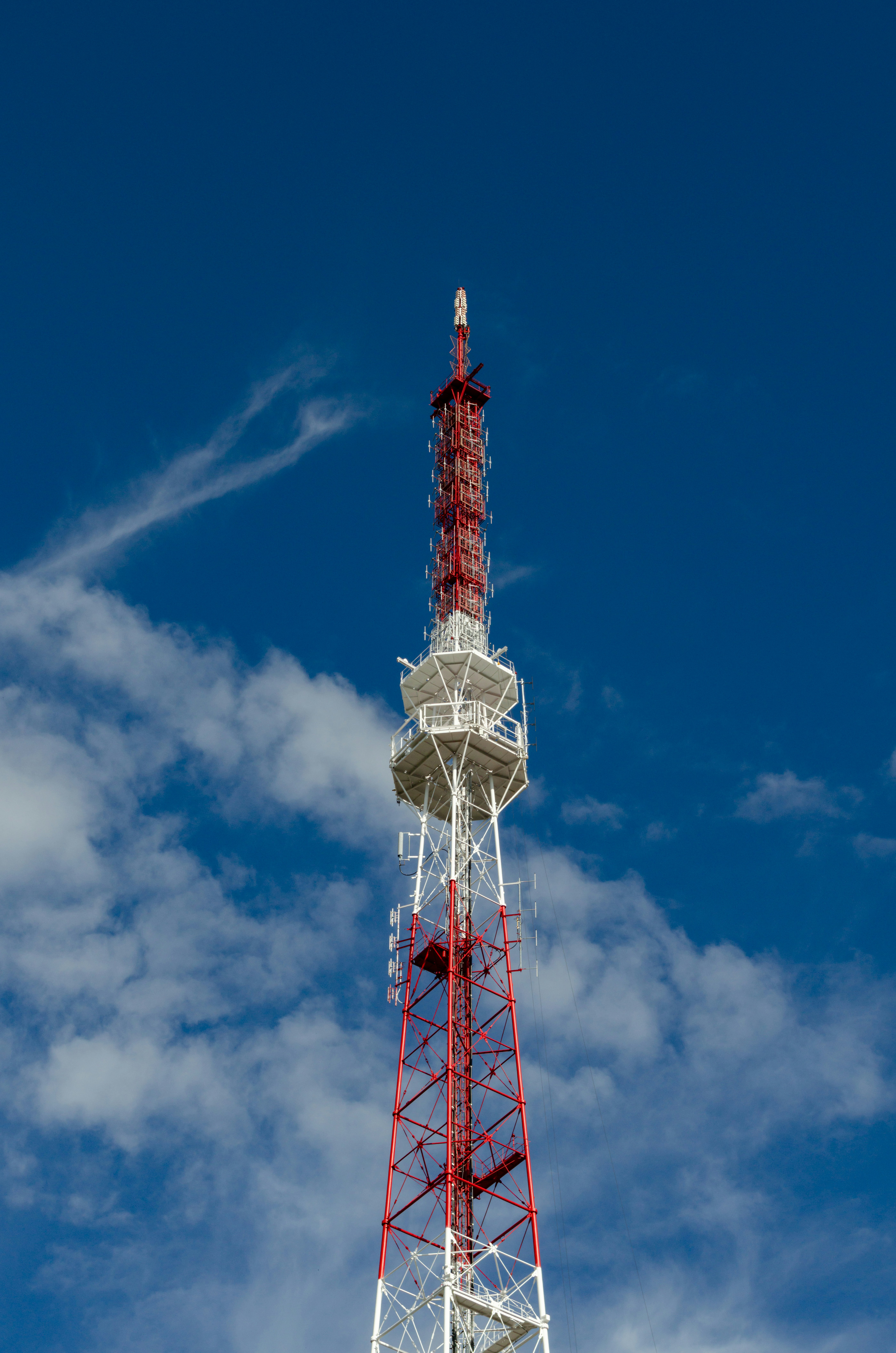 Tall communications tower rises into a clear blue sky with white clouds; a lattice structure transitioning from white to red at the upper sections, featuring an observation deck and antenna array. This is a structural photograph of a utility tower.