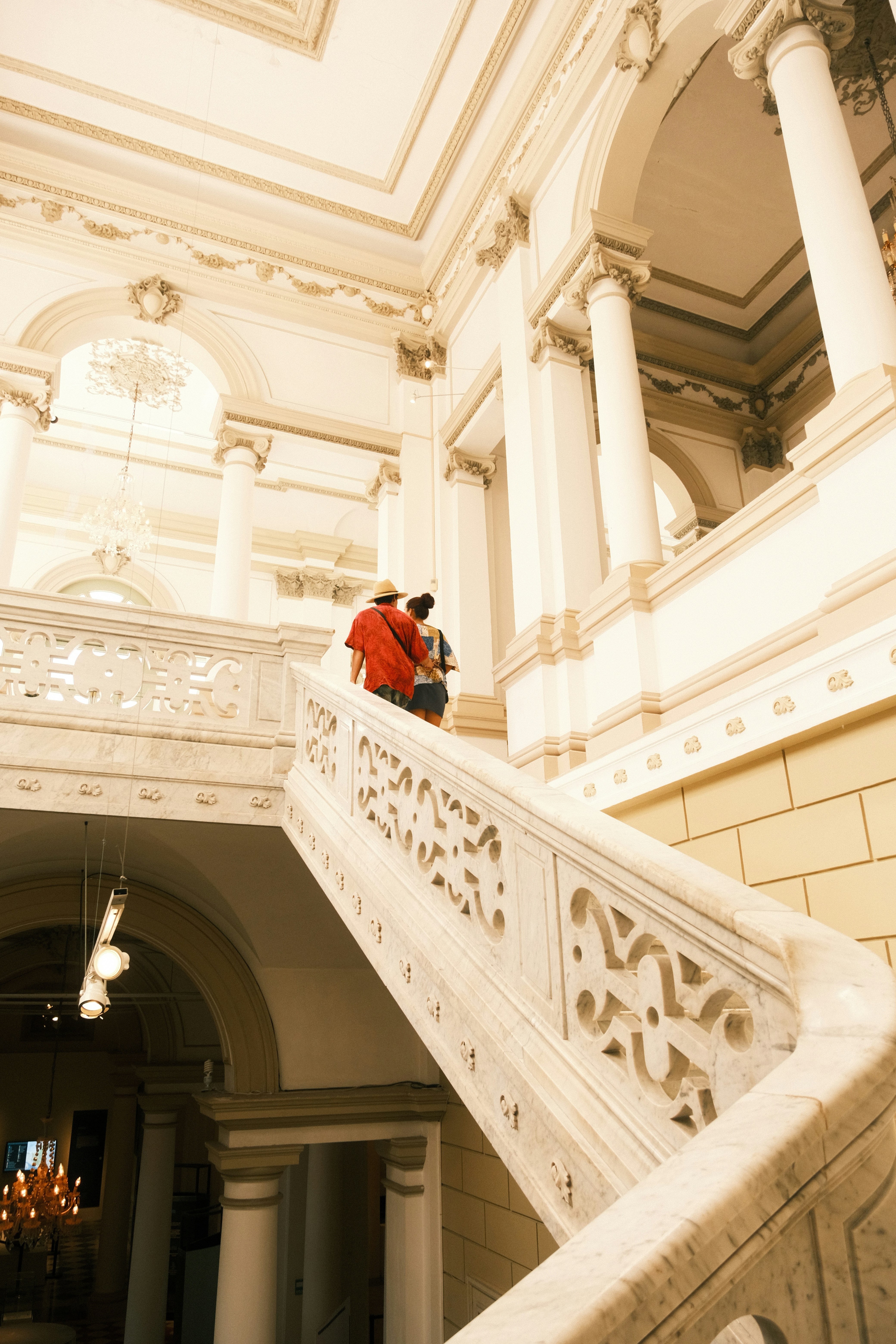 Two Tourists Ascend the Ornate Marble Staircase in a Sunlit Historic Hall