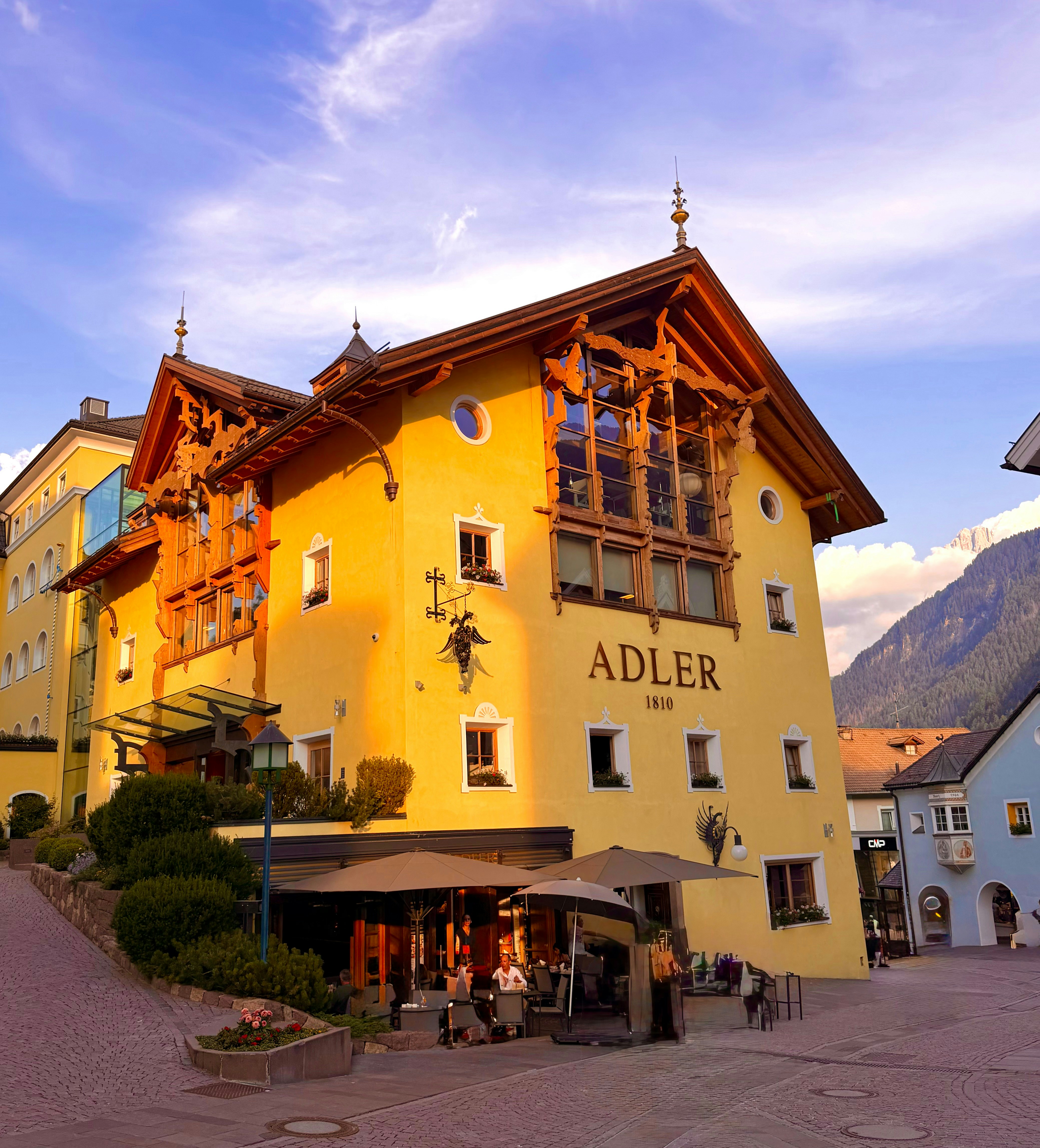 A sunlit yellow Alpine building with intricate wooden balconies, labeled 'ADLER', hosts an outdoor café beneath a clear sky; surrounding shops and mountains frame the scene.