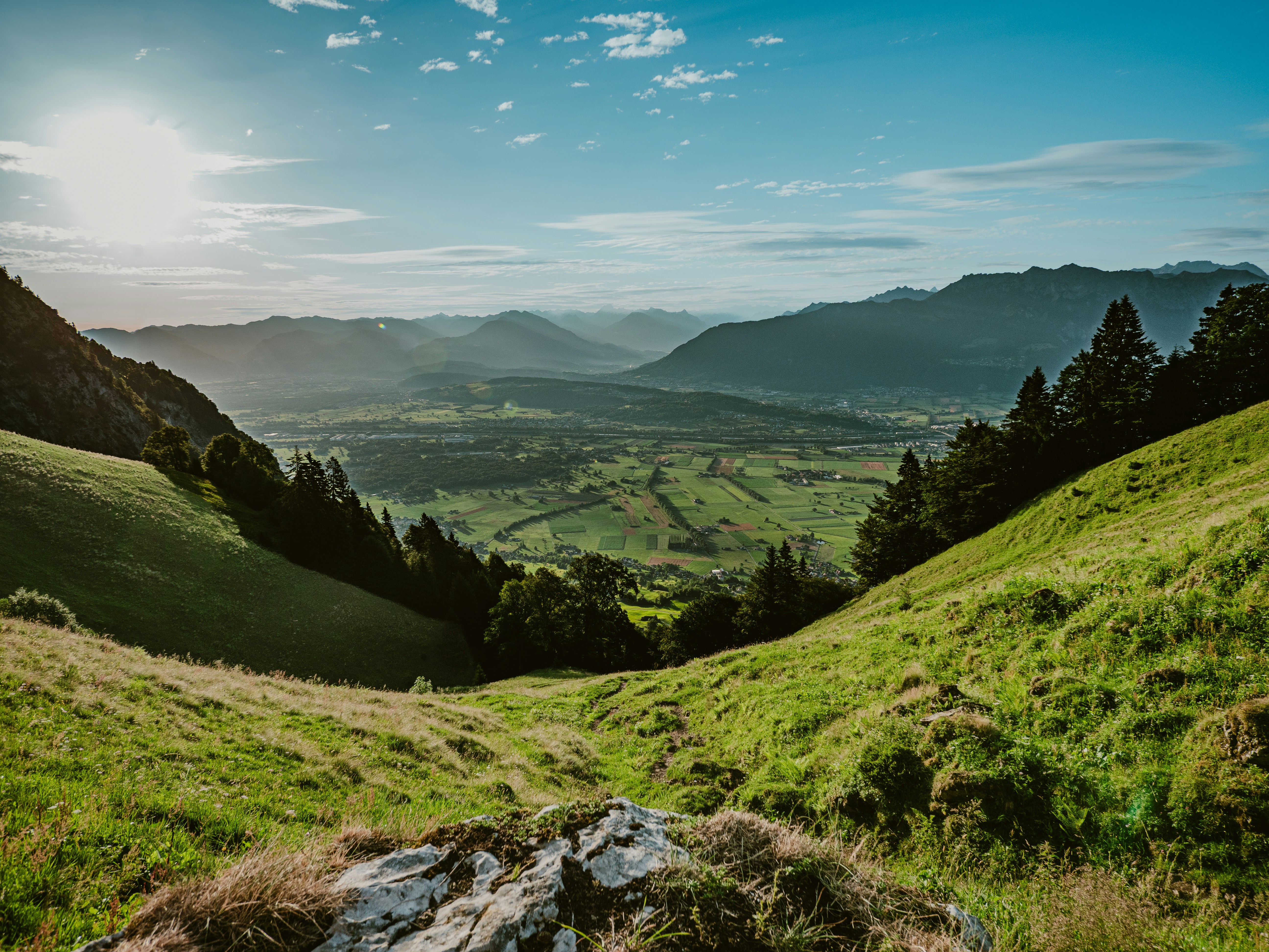 Sunlit valley unfolds from a grassy ridge beneath distant peaks