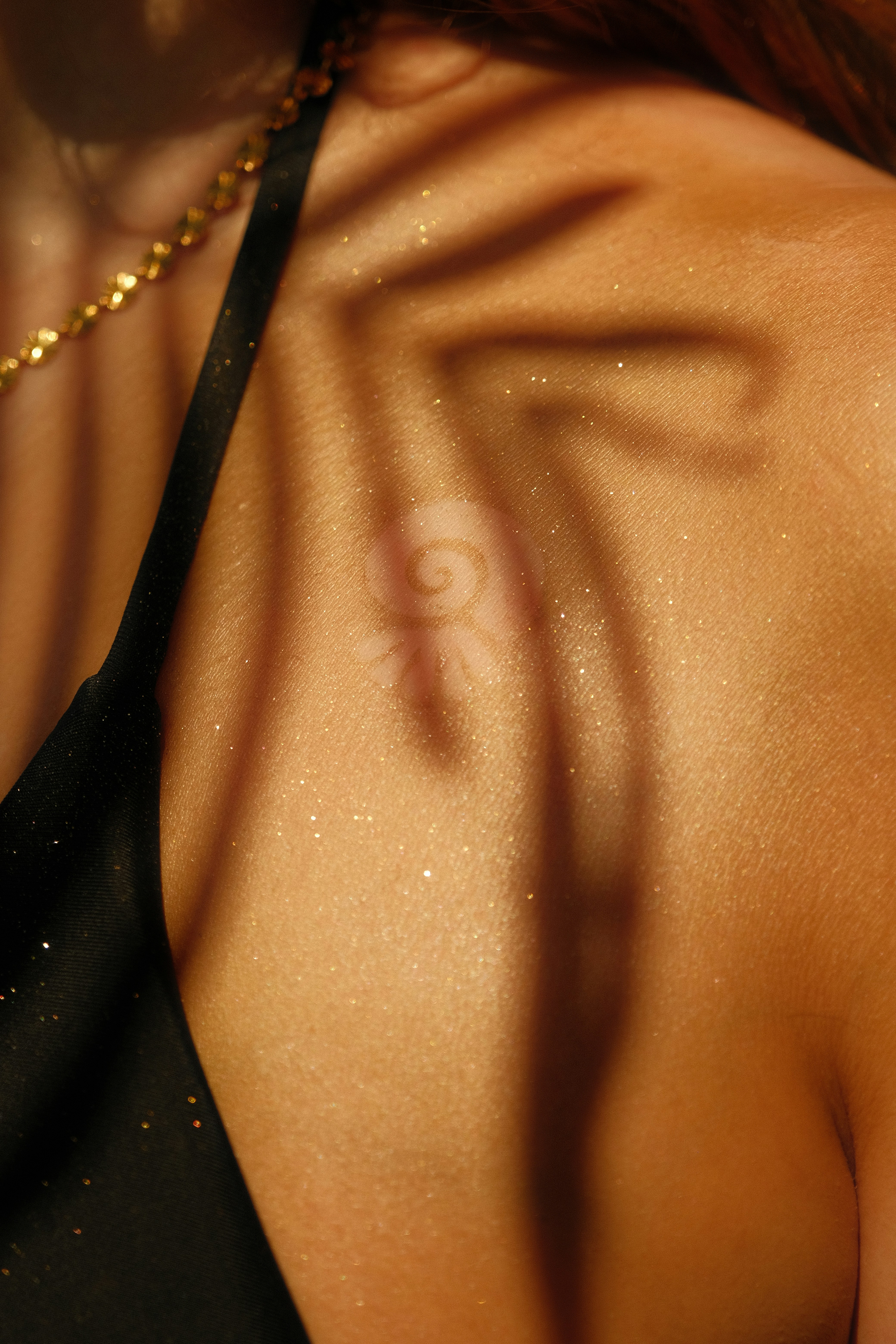 Glittering skin with a spiral shadow cast across the shoulder in a black bikini strap. A delicate gold chain rests near the edge of the frame, suggesting a close-up fashion or portrait shot.