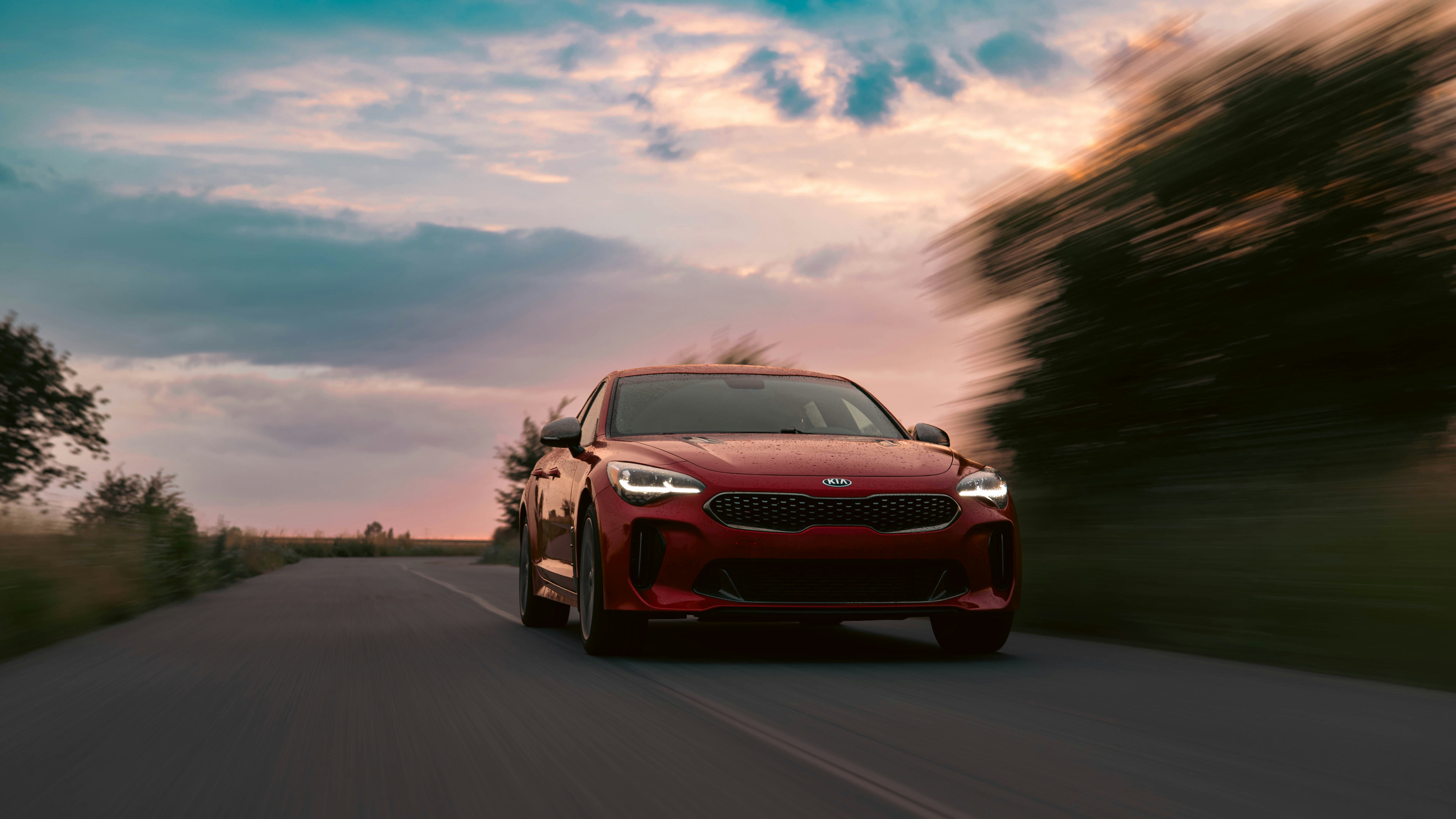 Red sports sedan speeding along a rural road with motion blur, low angle emphasizing front grille and headlights; no people visible, vehicle as main subject.