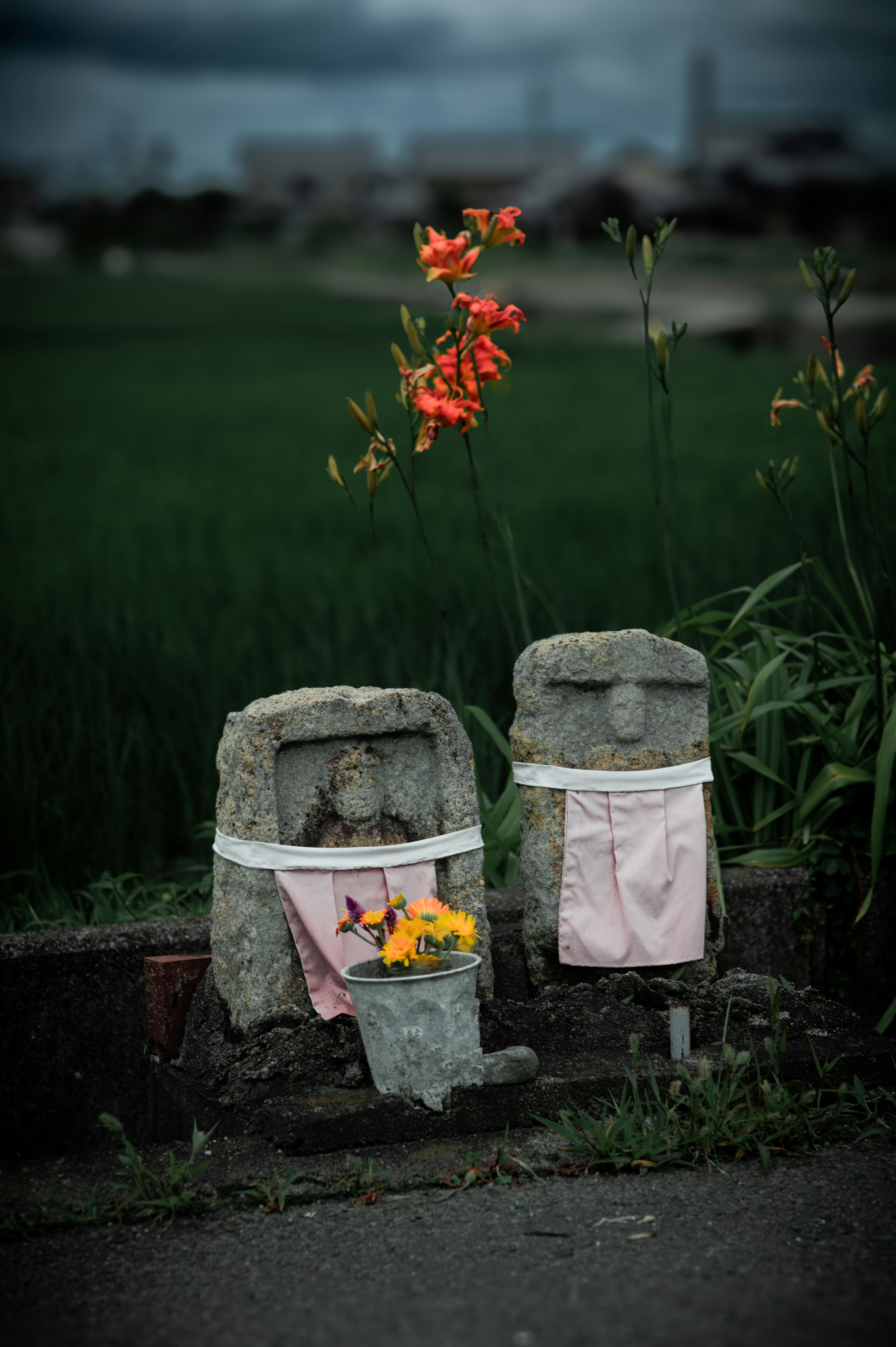 Two weathered stone markers with pink fabric bags sit beside a small metal bucket of yellow and purple blooms; a grassy field and distant industrial silhouettes form a muted background.