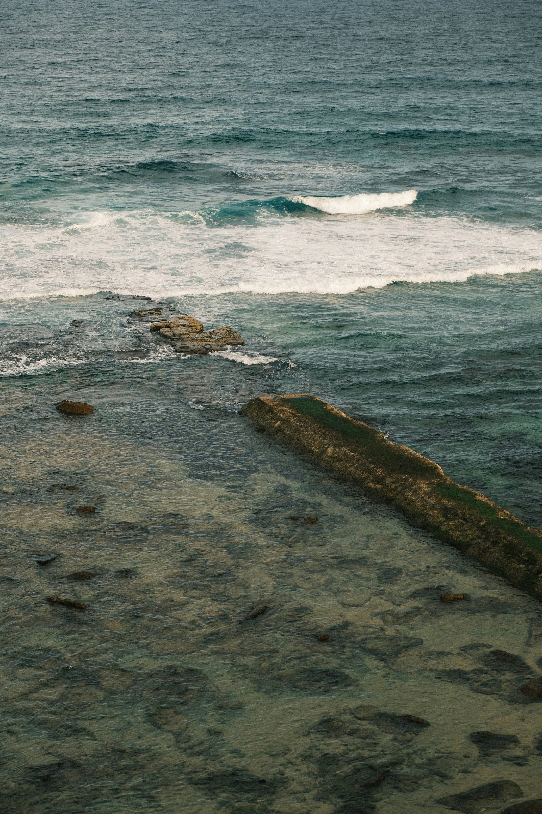 Clear coastal scene with a weathered jetty extending into shallow turquoise waters and gentle surf along a rocky shoreline.