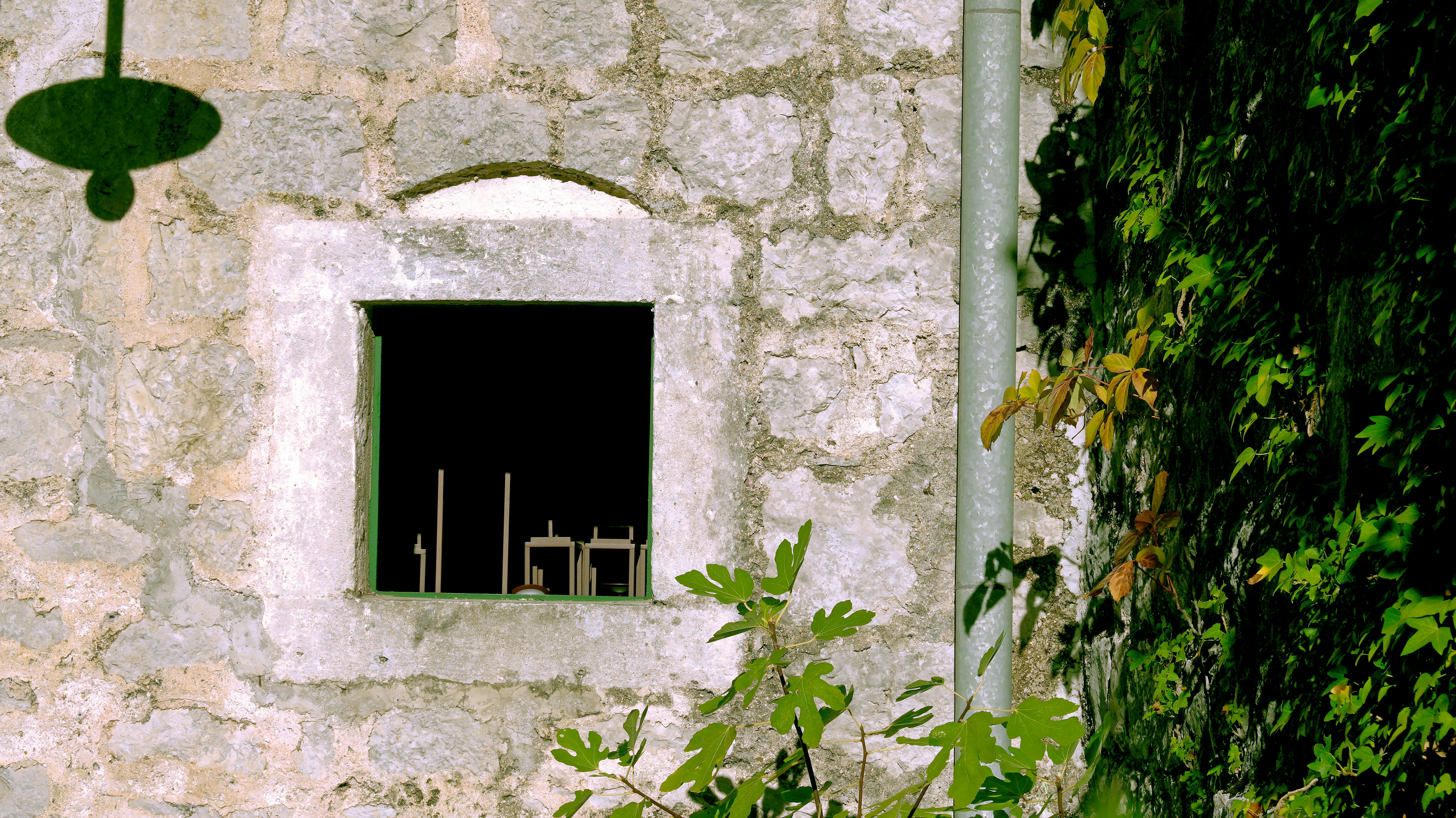 Crumbled stone wall with a dark square window and a metal pipe beside overgrown ivy, suggesting an abandoned or neglected building.