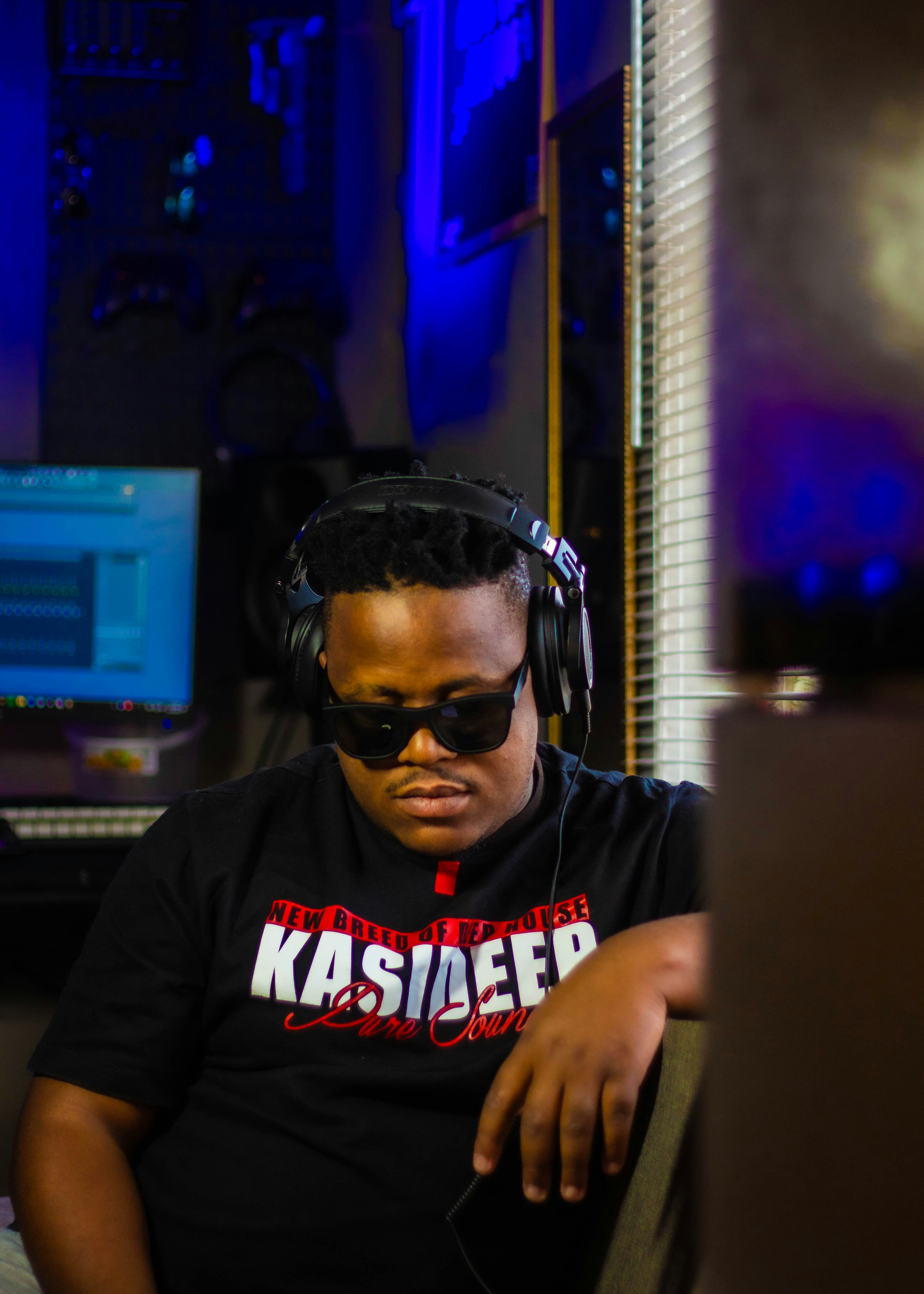Young man wearing sunglasses and headphones sits in a dimly lit studio with neon blue lighting, focusing on a music setup in the background. Logoed t-shirt and relaxed posture indicate a casual recording session.