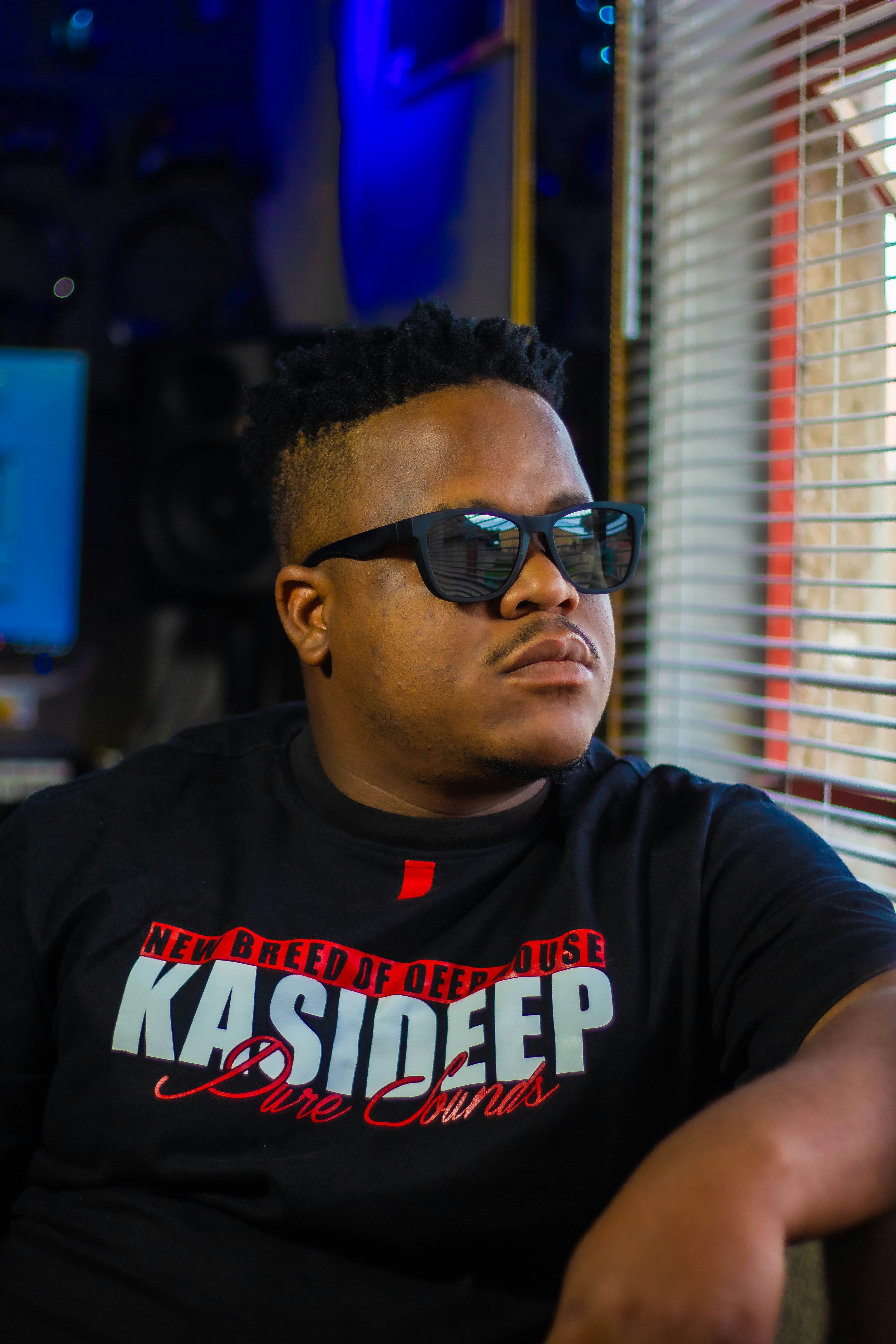 Dark-toned portrait of a man wearing sunglasses and a branded shirt, seated indoors with neon lighting and window blinds in the background. A candid, moody head-and-shoulders shot intended for social media branding.