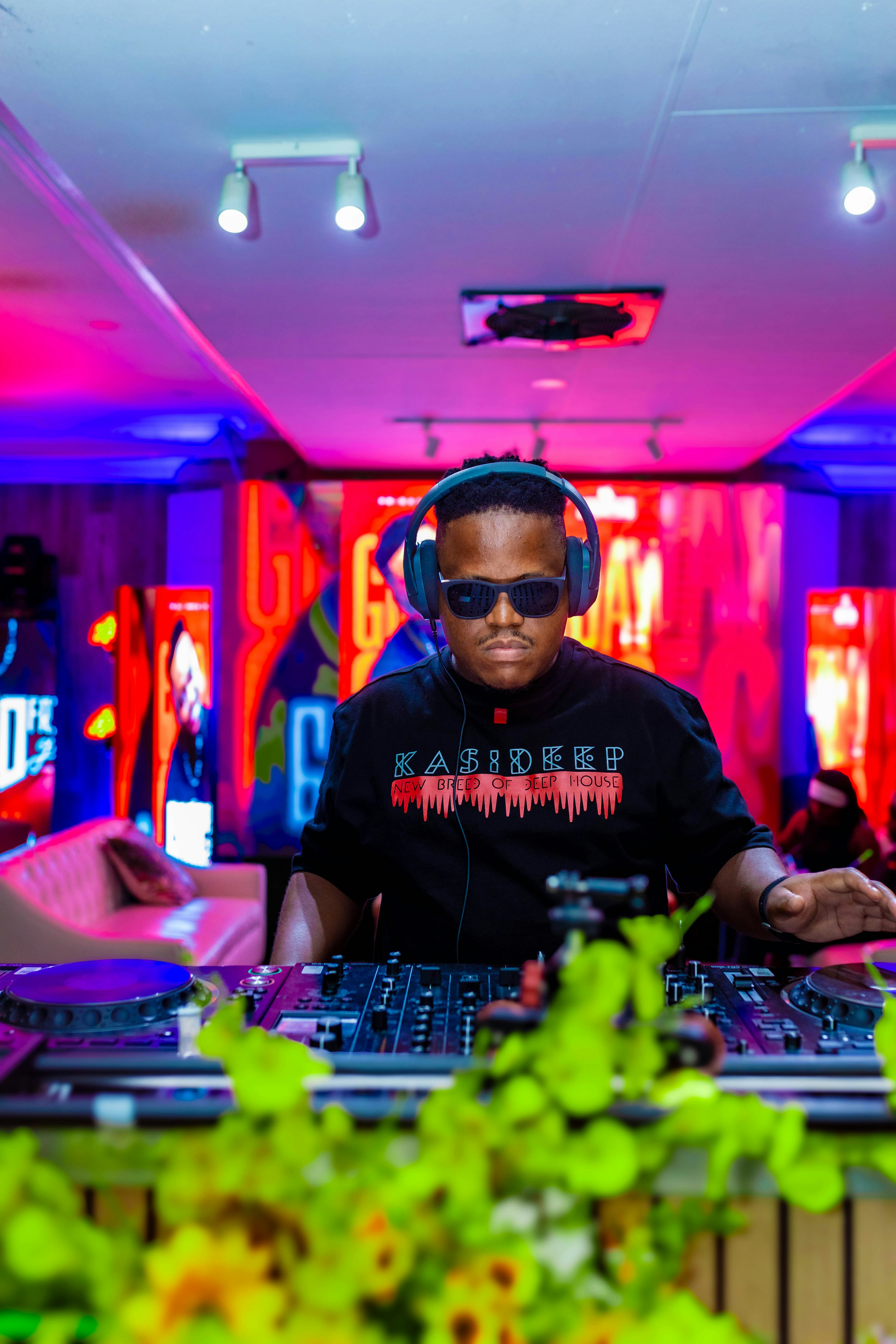 A DJ with headphones and sunglasses works the mixer in a neon-lit lounge; vibrant colors and plants frame the foreground, signaling a lively, modern club scene.
