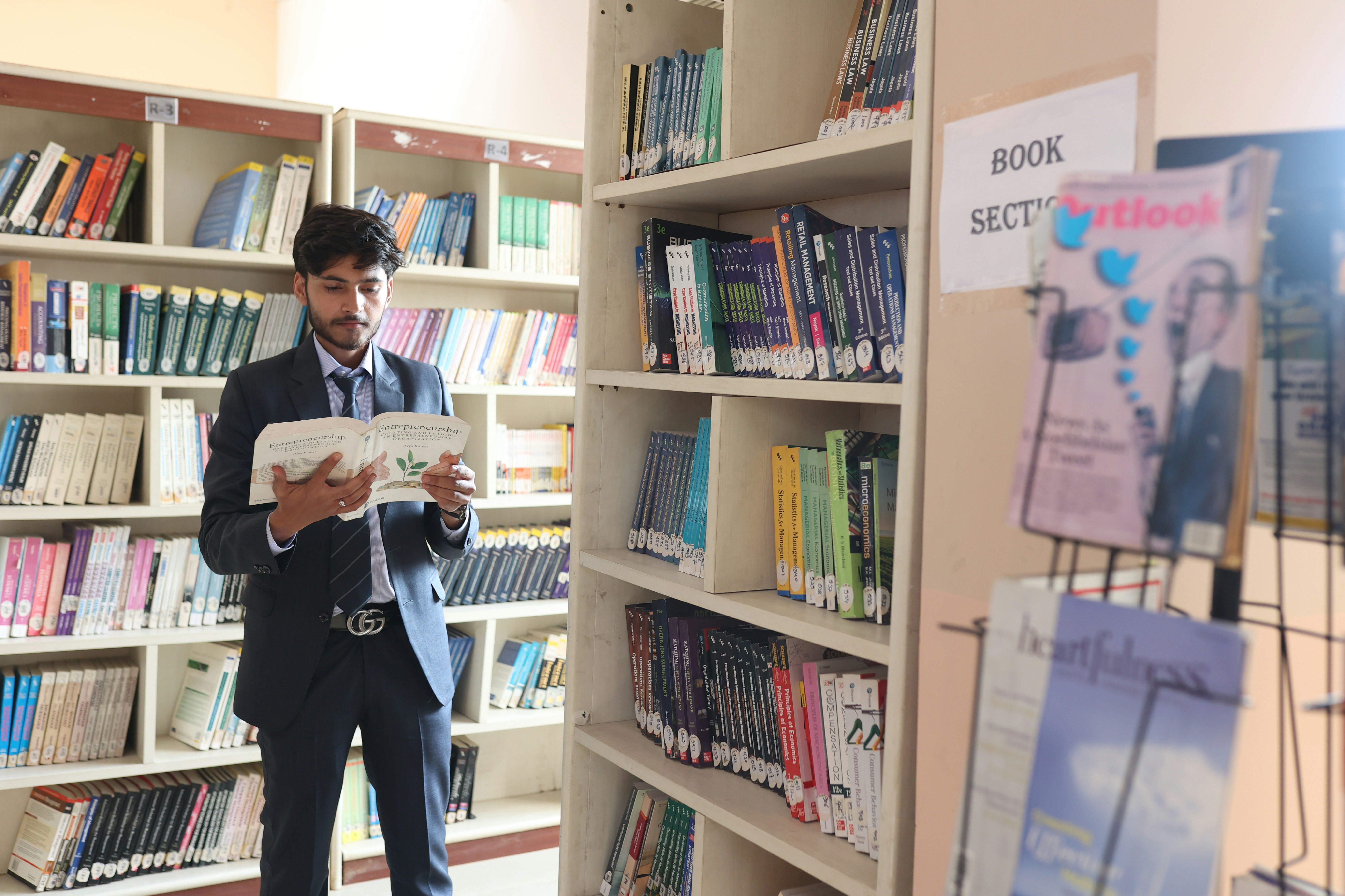 A man in a suit reads a book in a library aisle, with rows of colorful books on shelves behind him. A magazine display appears on the right side, indicating a library or bookstore setting.